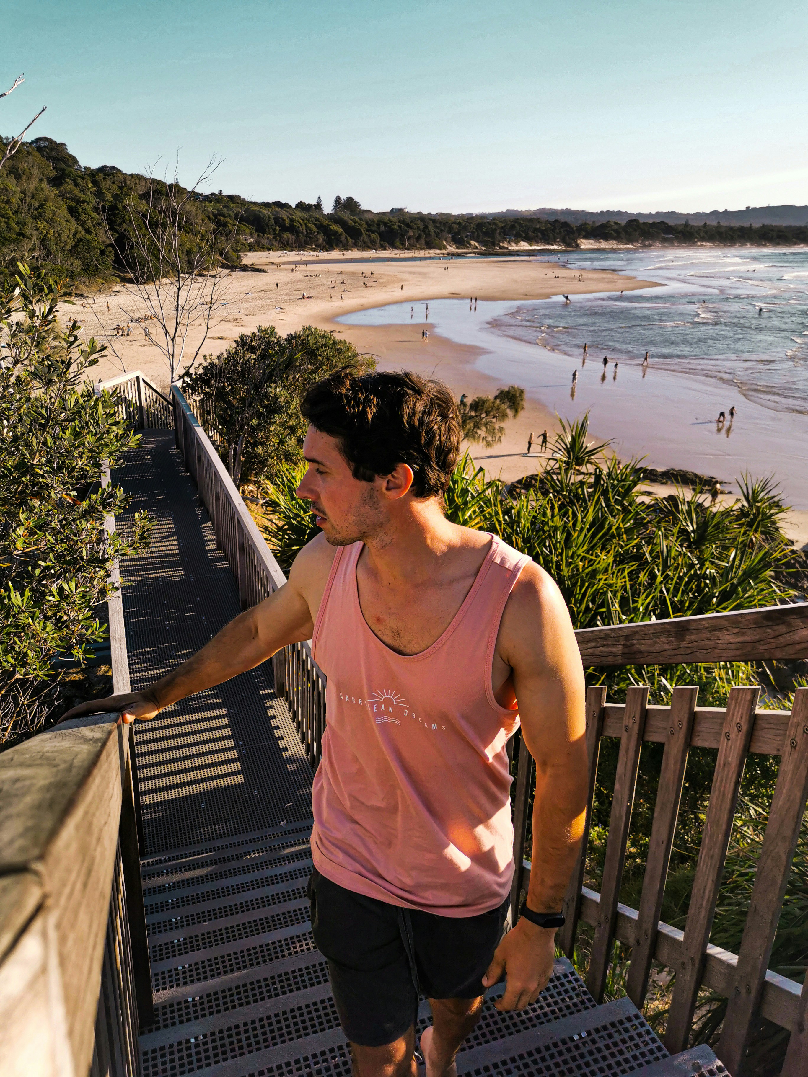 Sunlit ascent: a man in pink tank climbs a coastal boardwalk as the beach and sea stretch out below