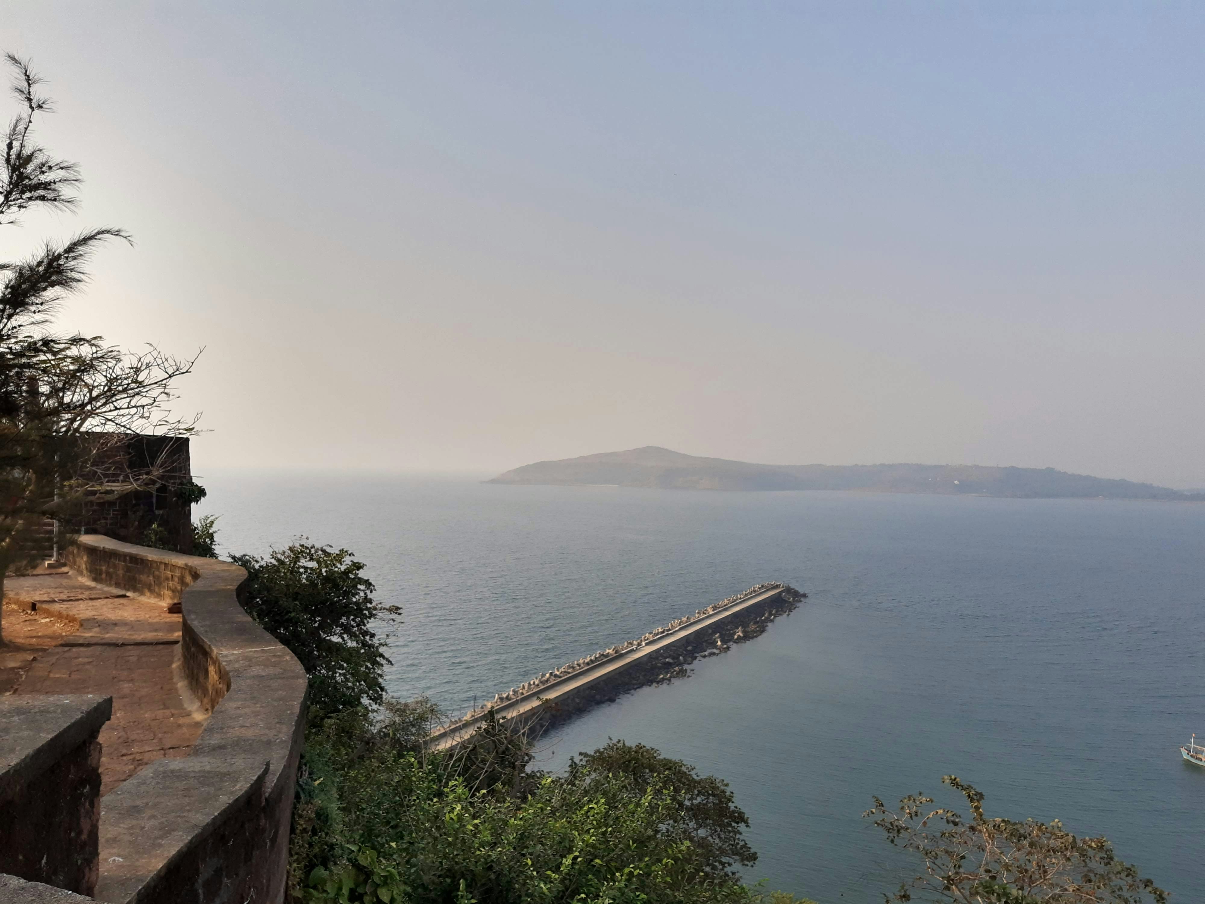 Serene coastal view featuring a jetty extending into the calm sea, framed by lush greenery and distant hills.