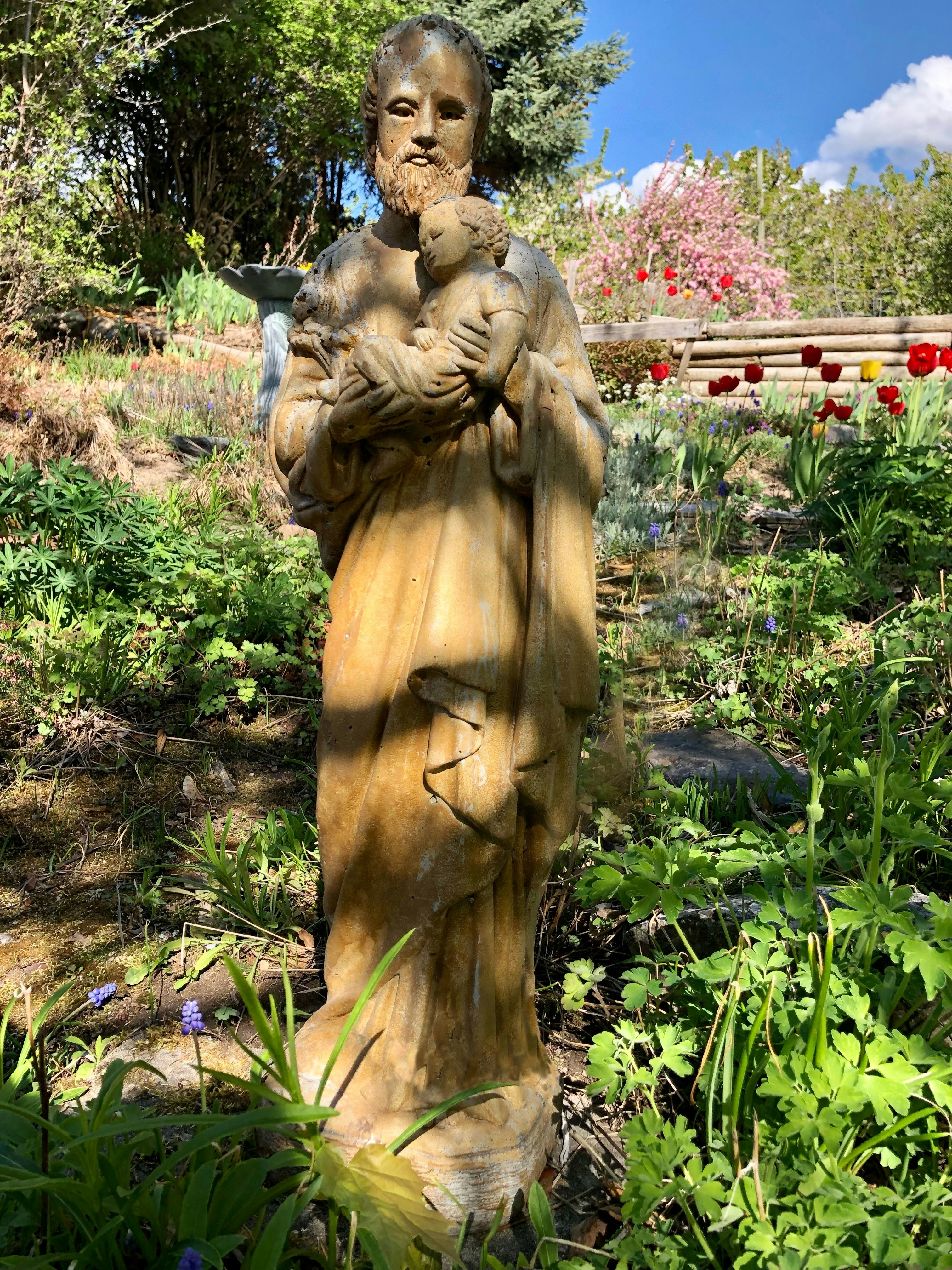 Weathered statue of a figure holding a child, surrounded by vibrant flowers in a lush garden setting.