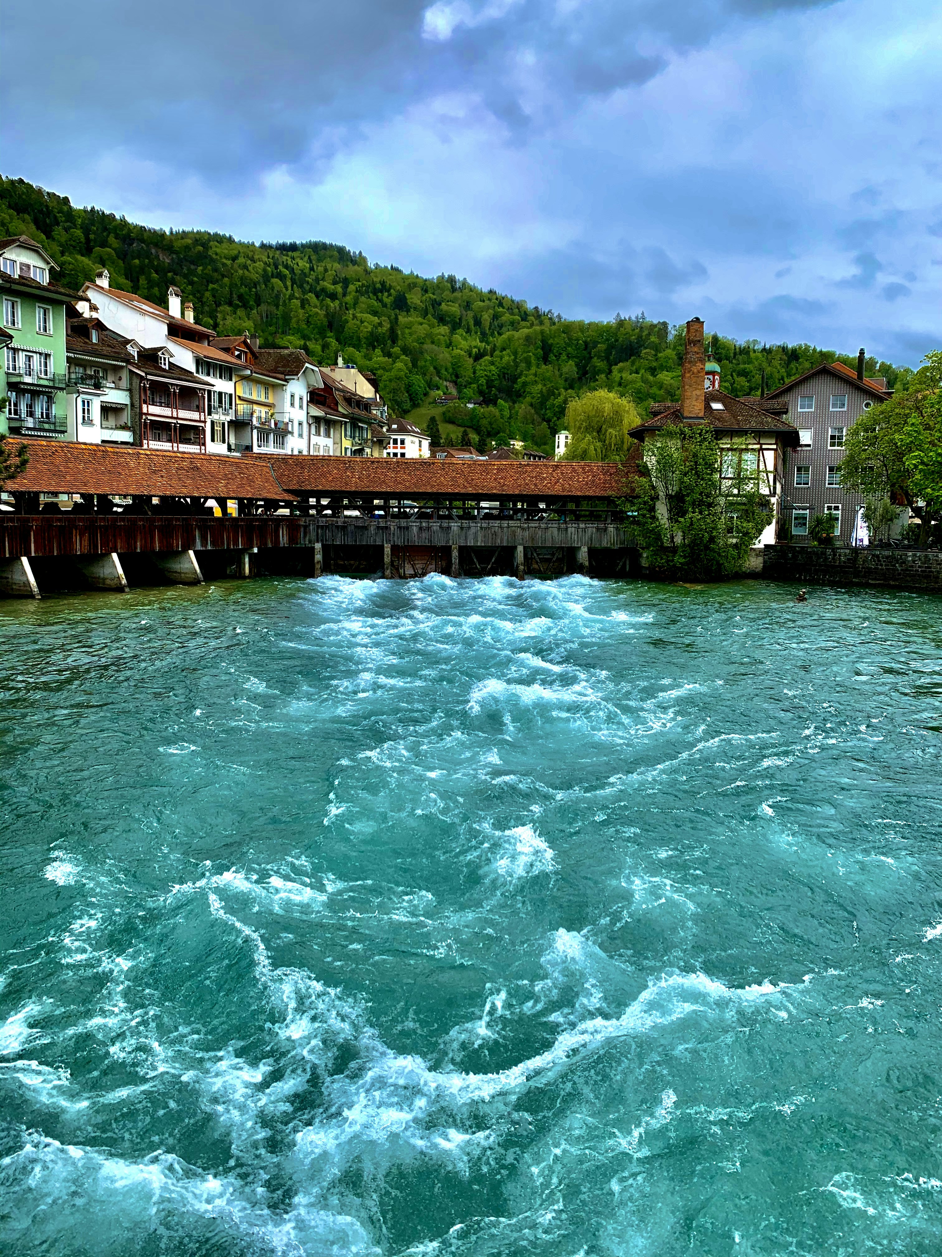 Vibrant turquoise river cascading past historic buildings under a moody sky, showcasing the harmony between nature and architecture.