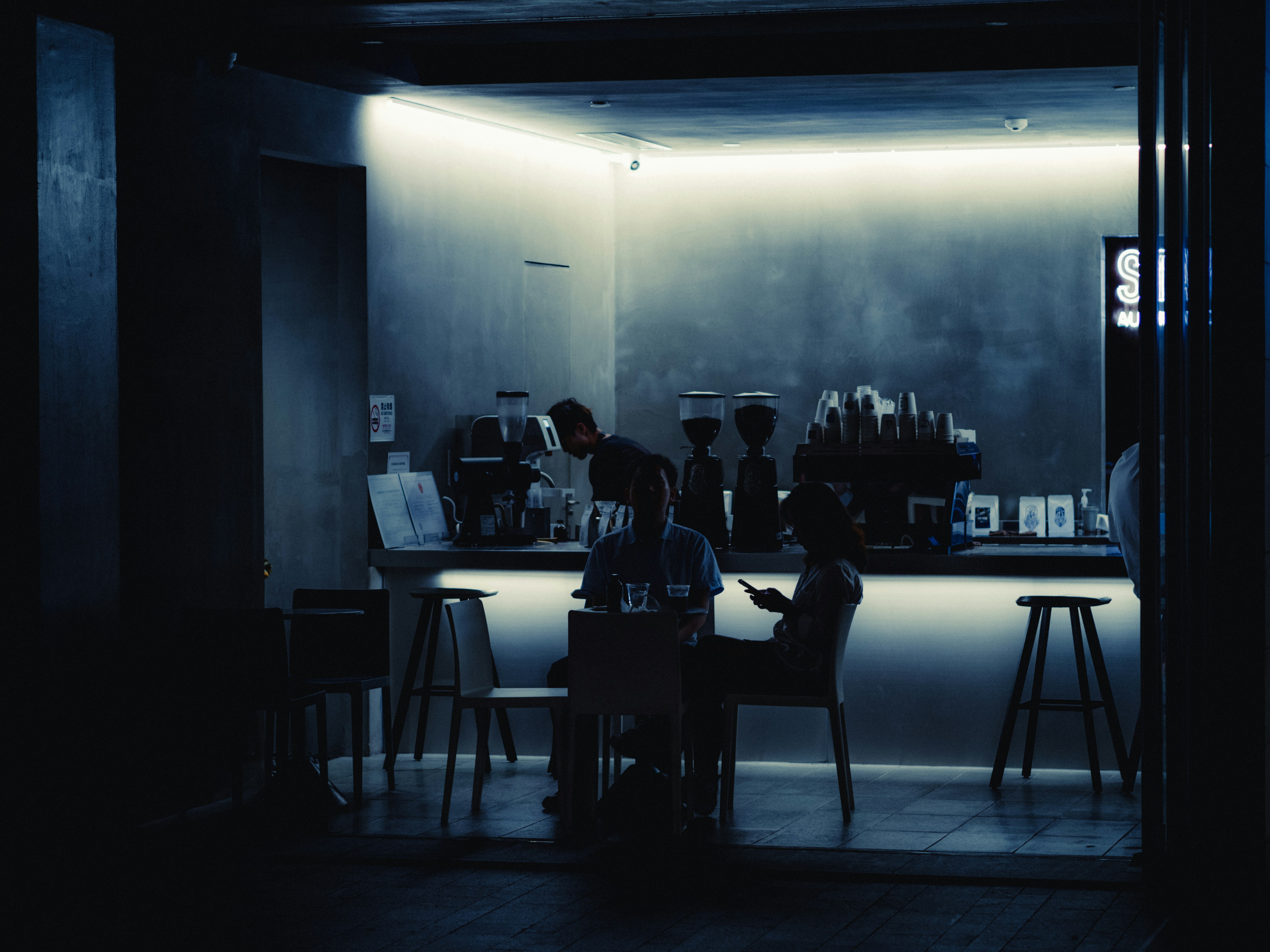 Dimly lit cafe interior bathed in blue neon light, with a small group seated at a table near the bar.