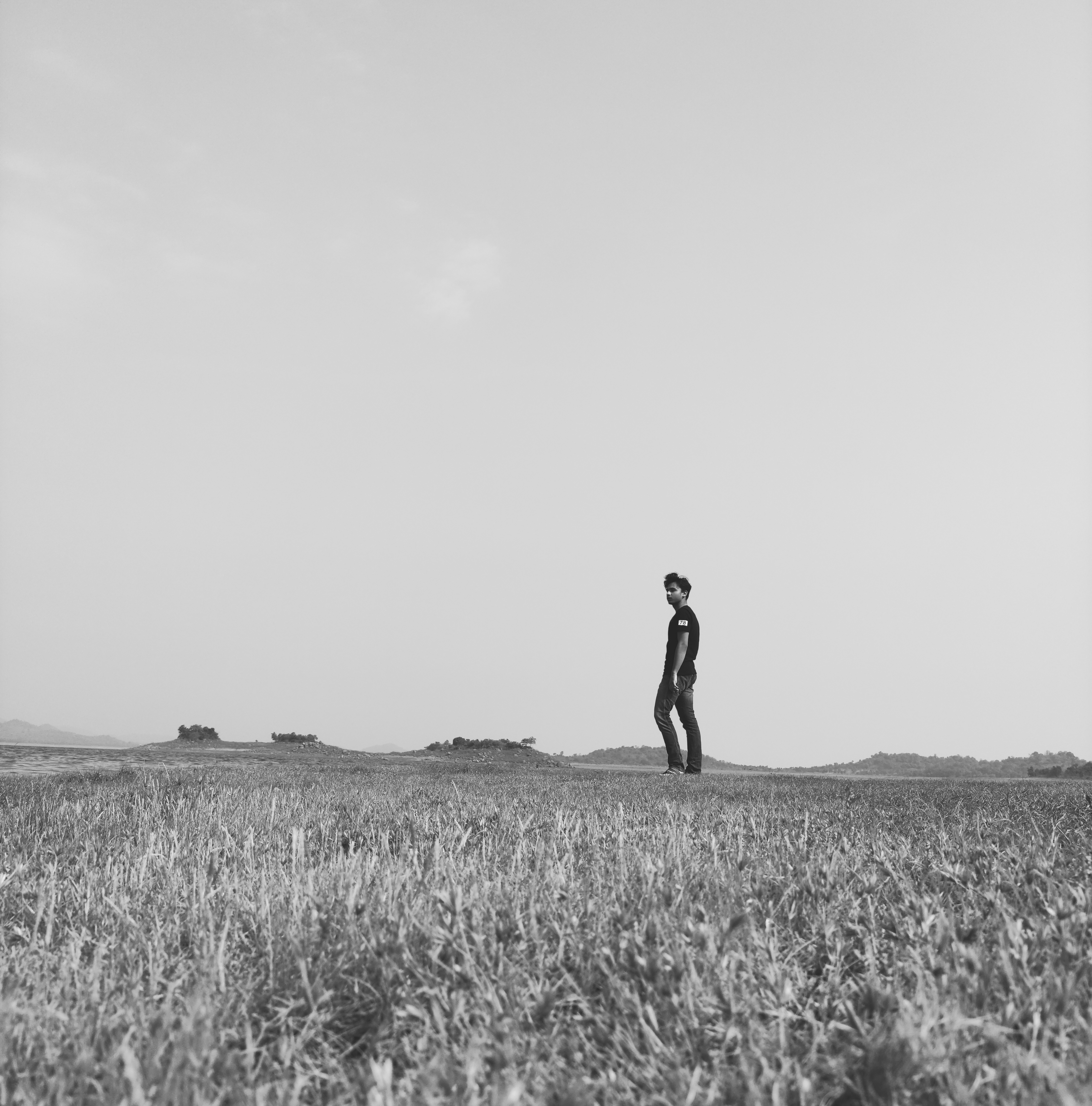 Monochrome photograph of a solitary figure standing in a wide grass field beneath a vast sky.