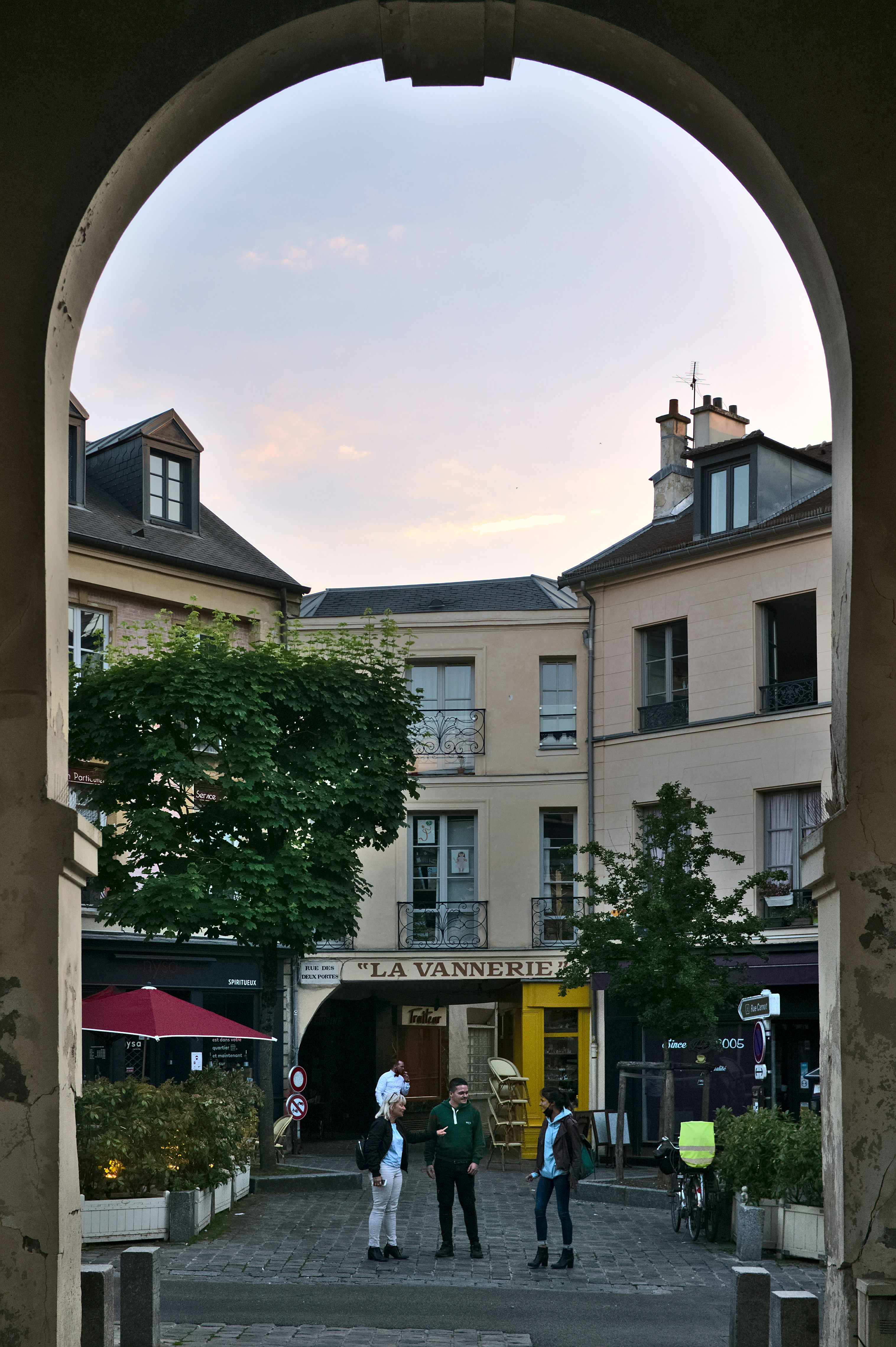 Three individuals engage in conversation beneath an archway, framed by charming Parisian architecture and a colorful evening sky.