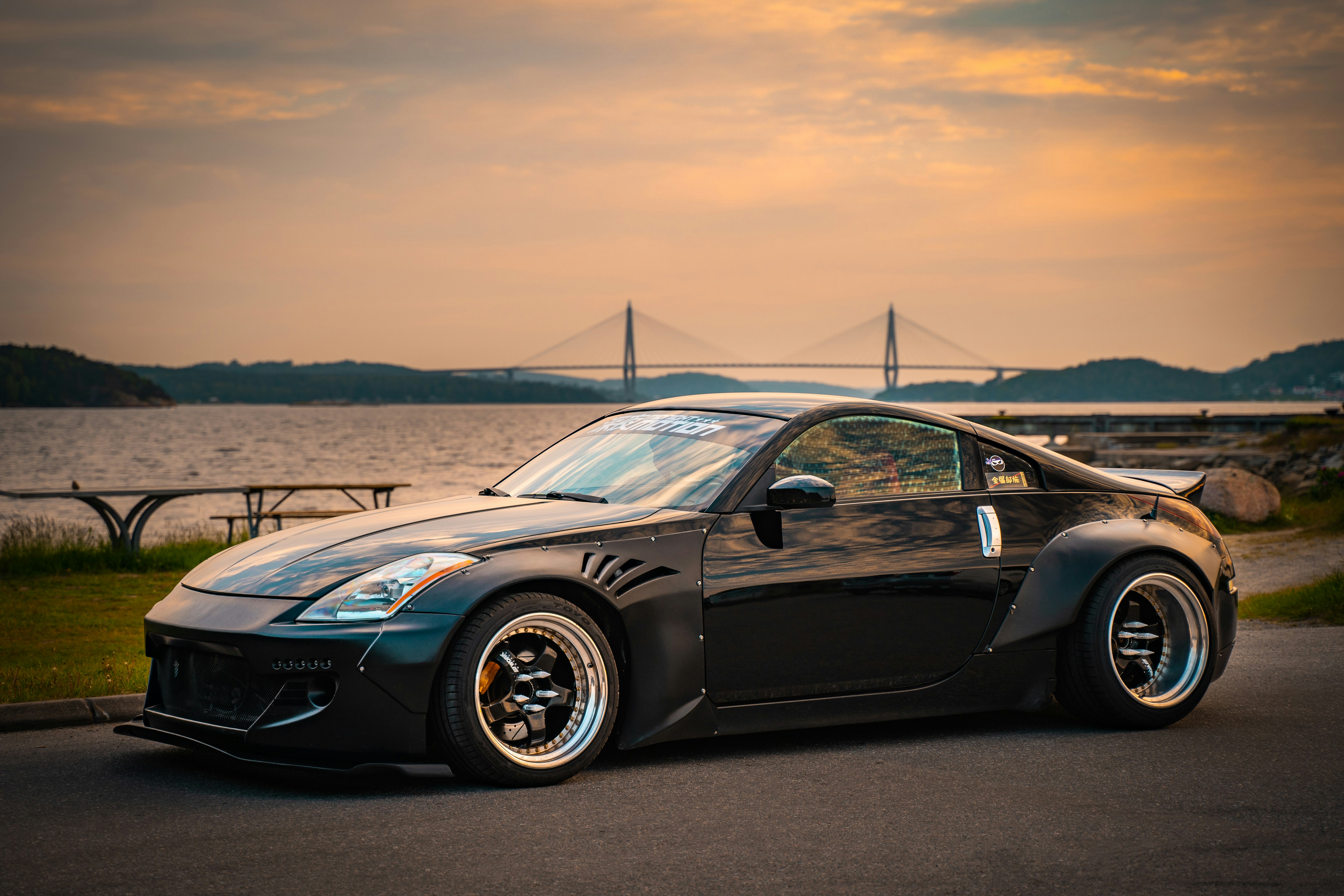 Black Porsche 911 parked on a road near a scenic waterfront with a bridge in the background at sunset.