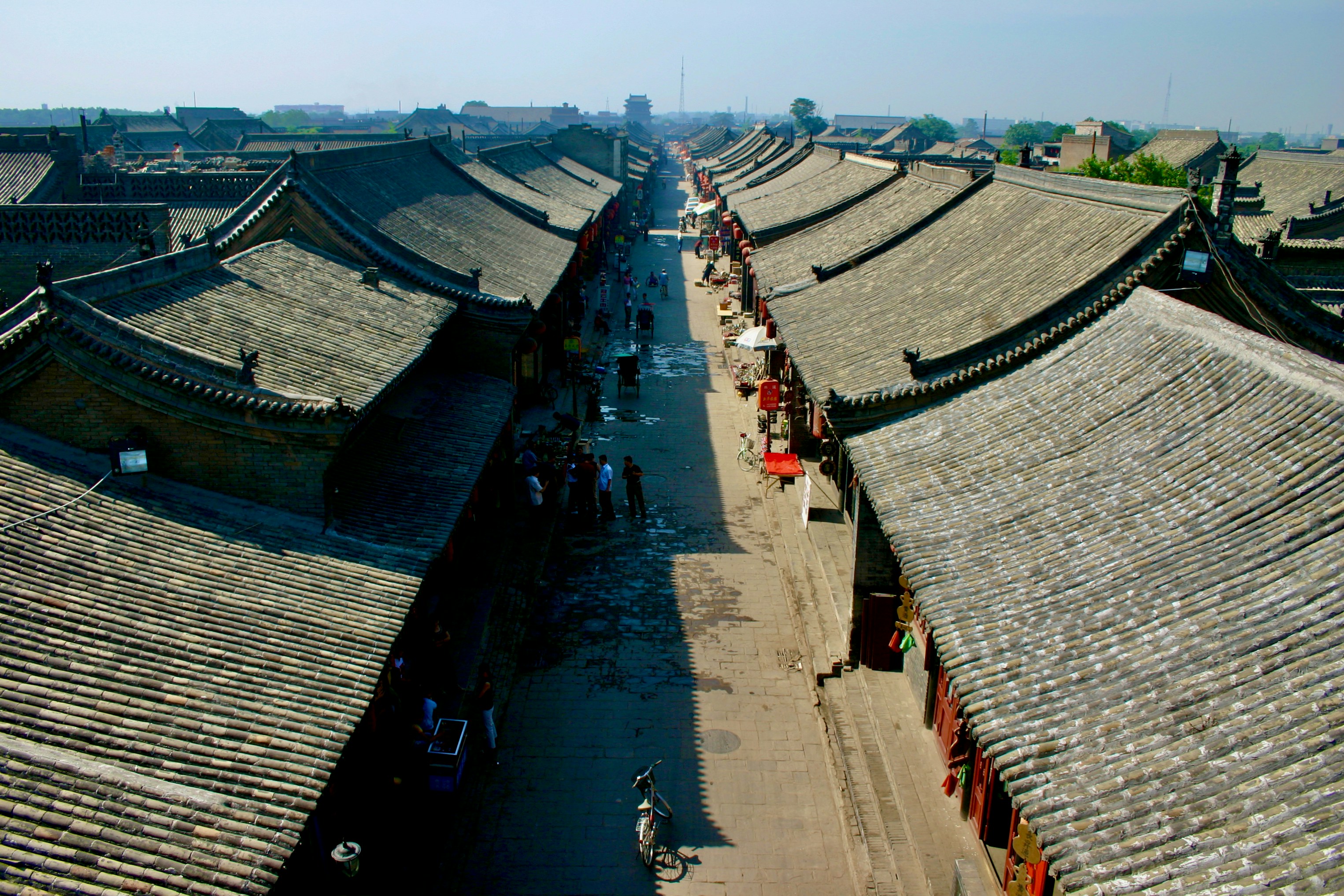 Aerial view of a historic market street lined with traditional rooftops, showcasing the vibrant atmosphere of daily life and commerce. Sunlight bathes the scene, revealing the textures of the aged architecture.