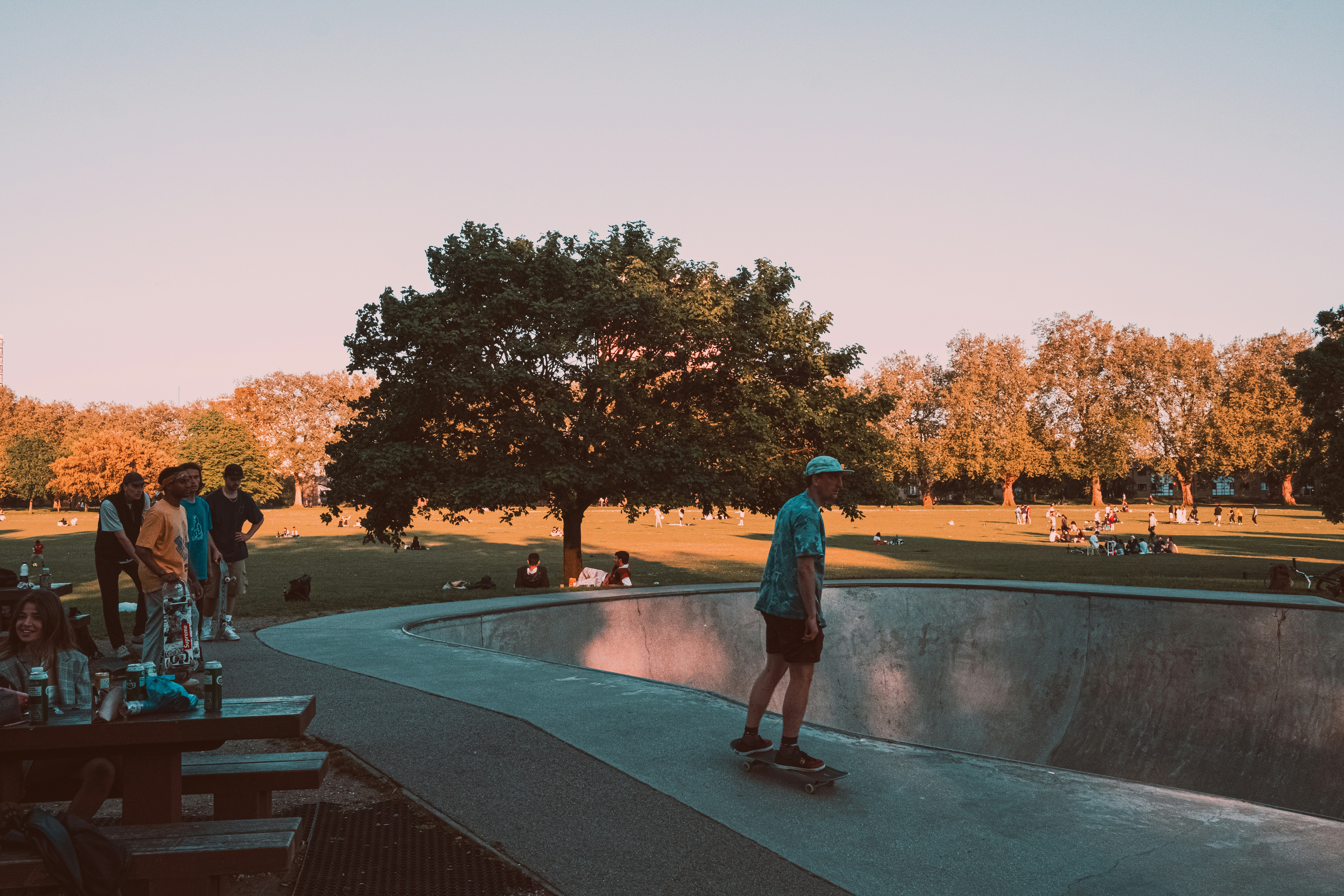 A skateboarder glides through a park skate bowl as spectators enjoy the warm evening light. The scene captures the relaxed vibe of an urban outdoor gathering.