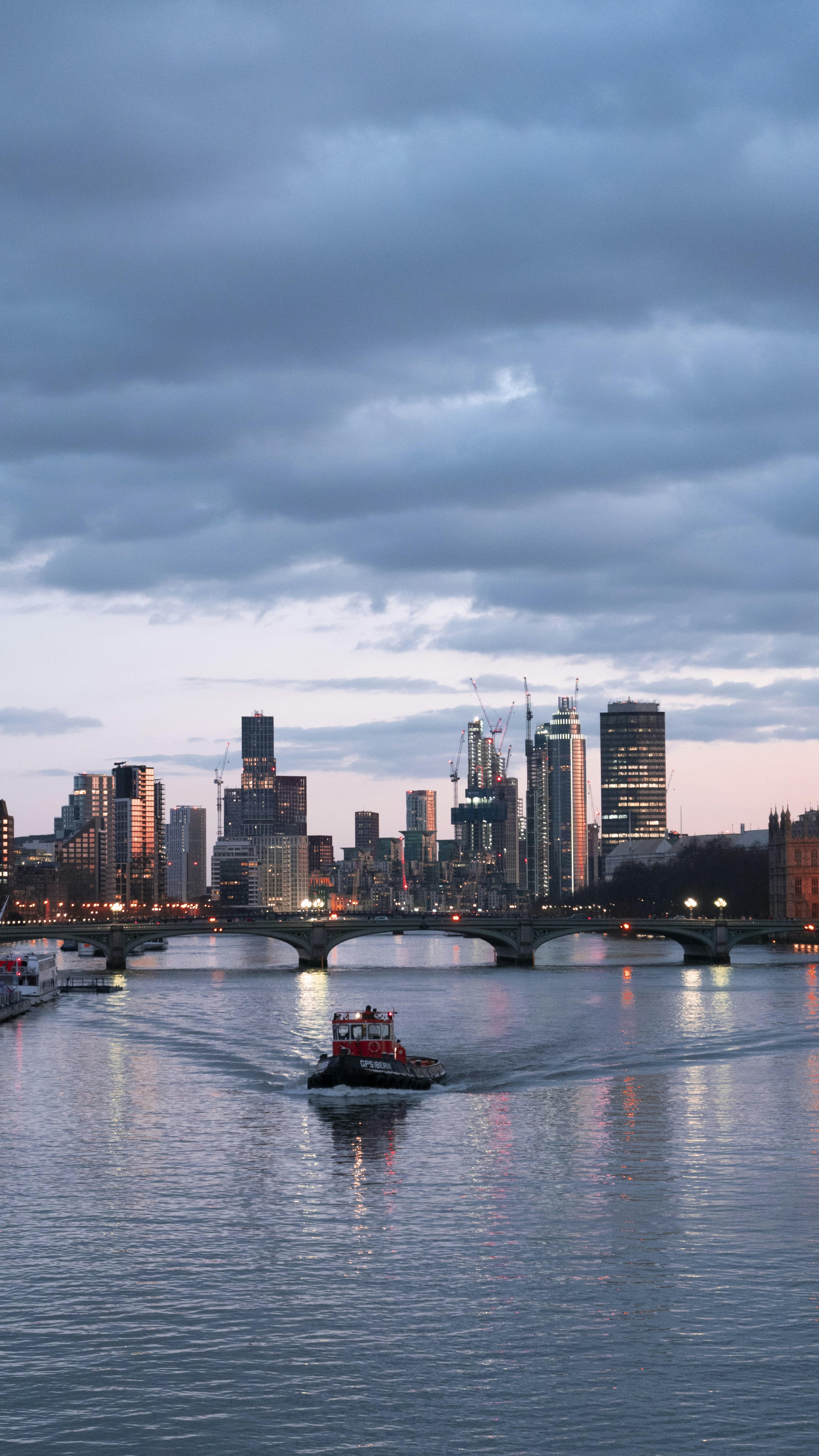 A boat navigates the calm waters of the Thames, framed by a skyline of modern skyscrapers under a twilight sky.