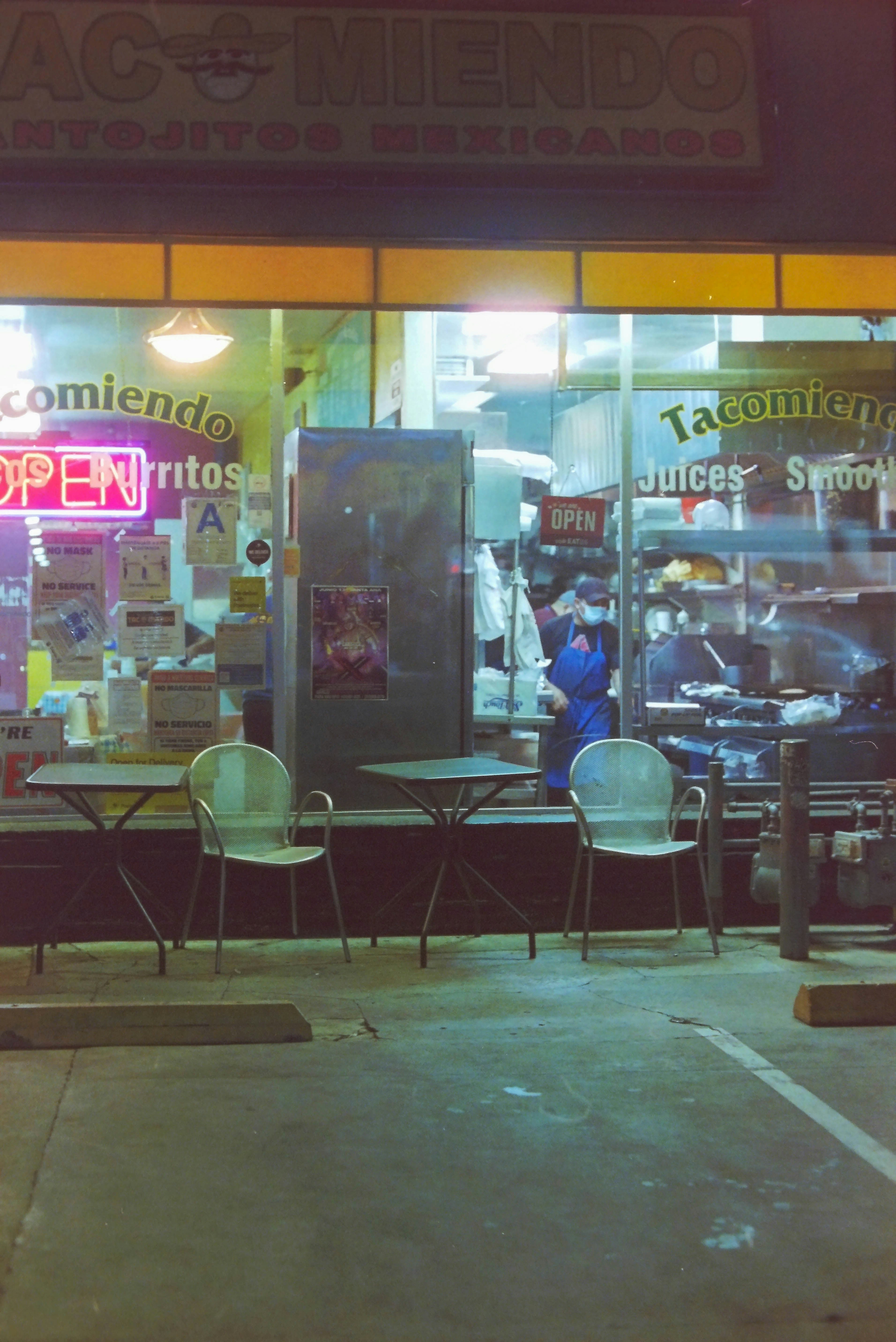 Nighttime photo of a taco shop window with neon Open sign and outdoor seating.