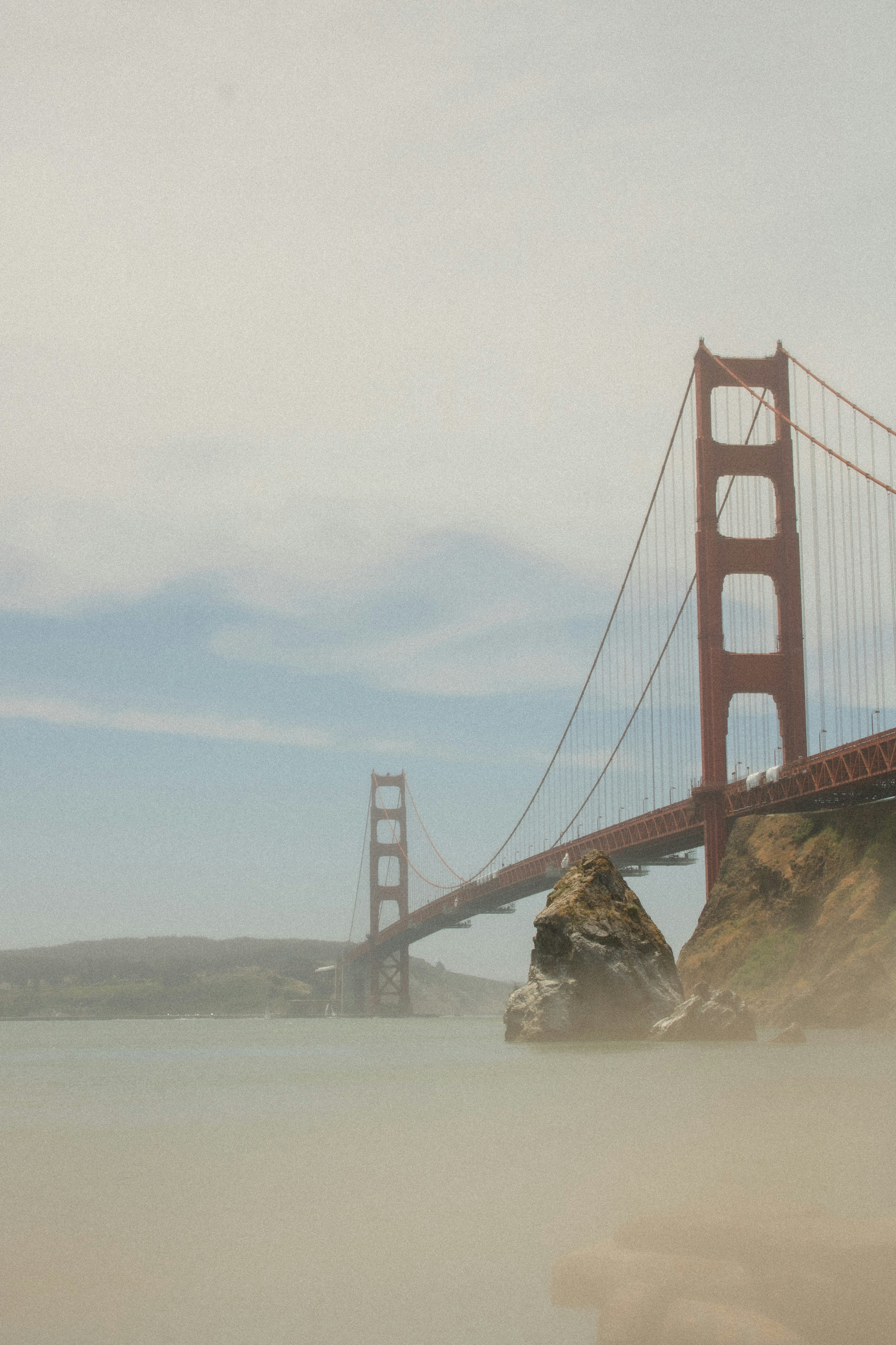 Golden Gate Bridge partially shrouded in fog, with rocky shoreline and calm waters in the foreground.
