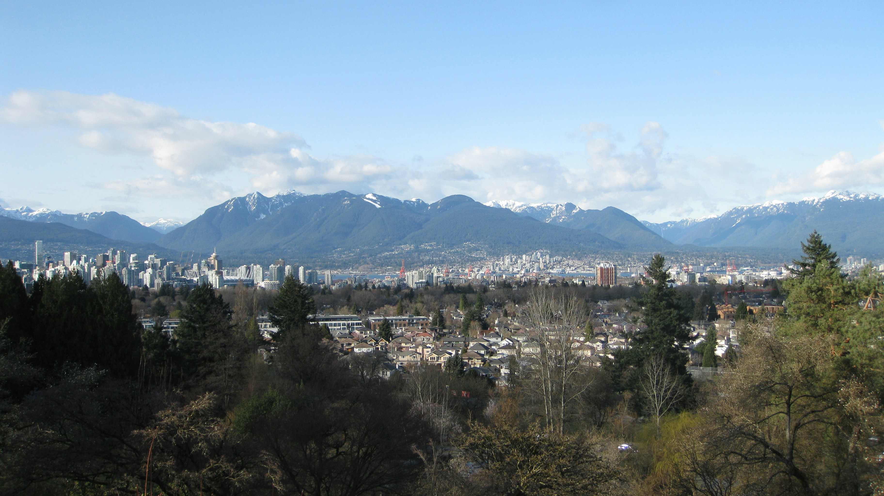 Cityscape framed by lush trees and expansive mountains under a clear blue sky.