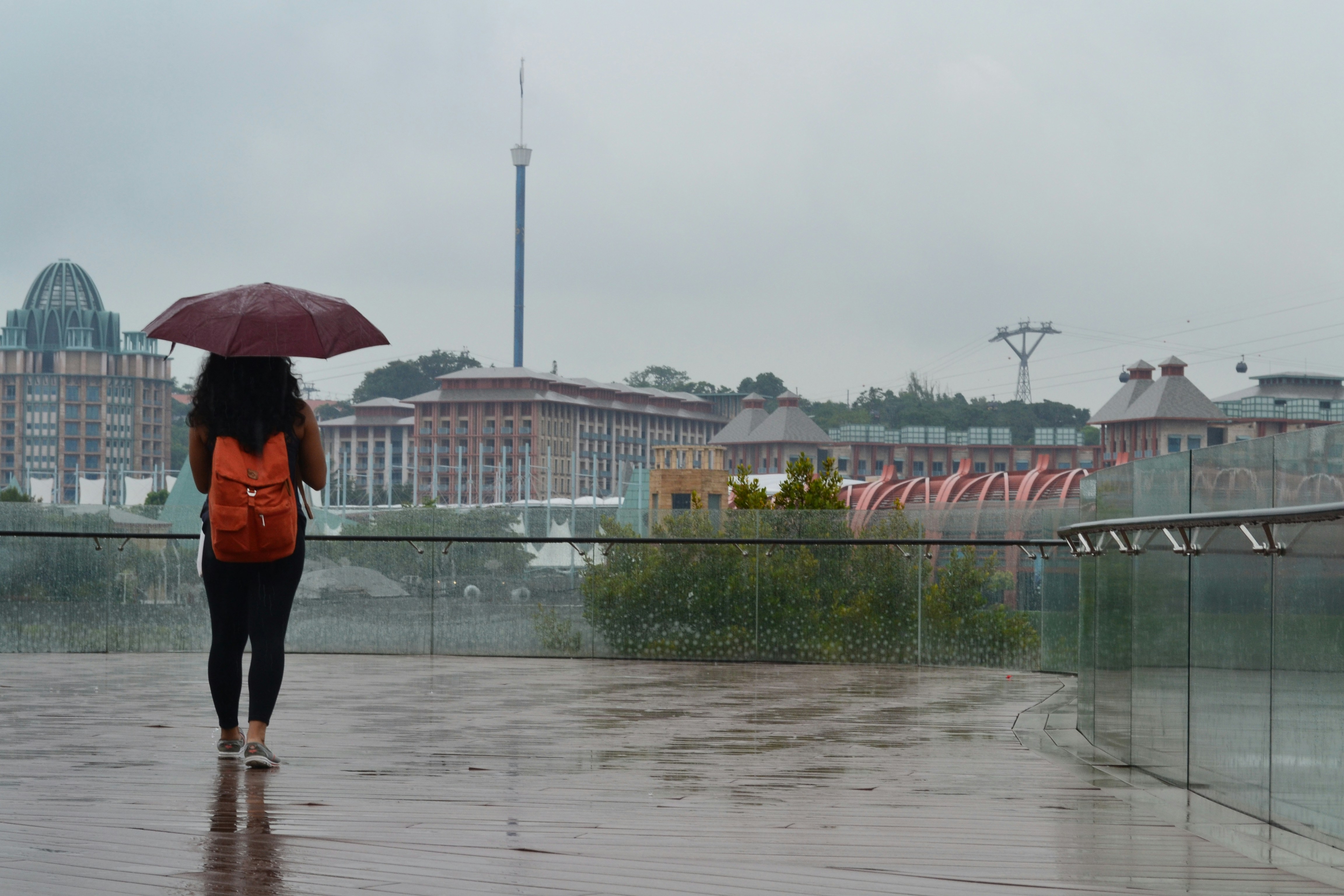 Person with an umbrella stands on a wet wooden deck, gazing at a distant urban landscape under overcast skies.