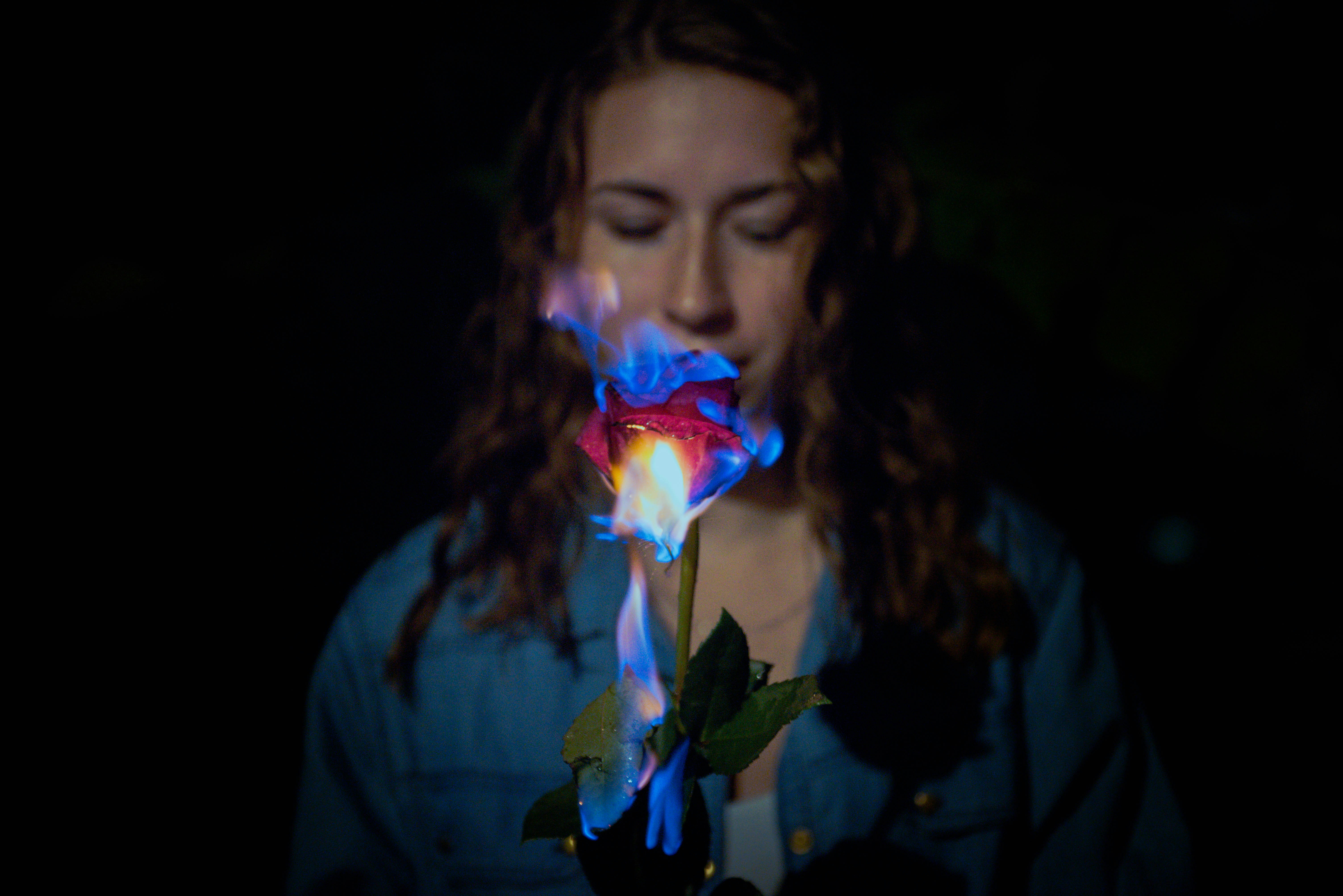 Woman holding a rose set ablaze, contrasting vibrant blue flames against a dark background.