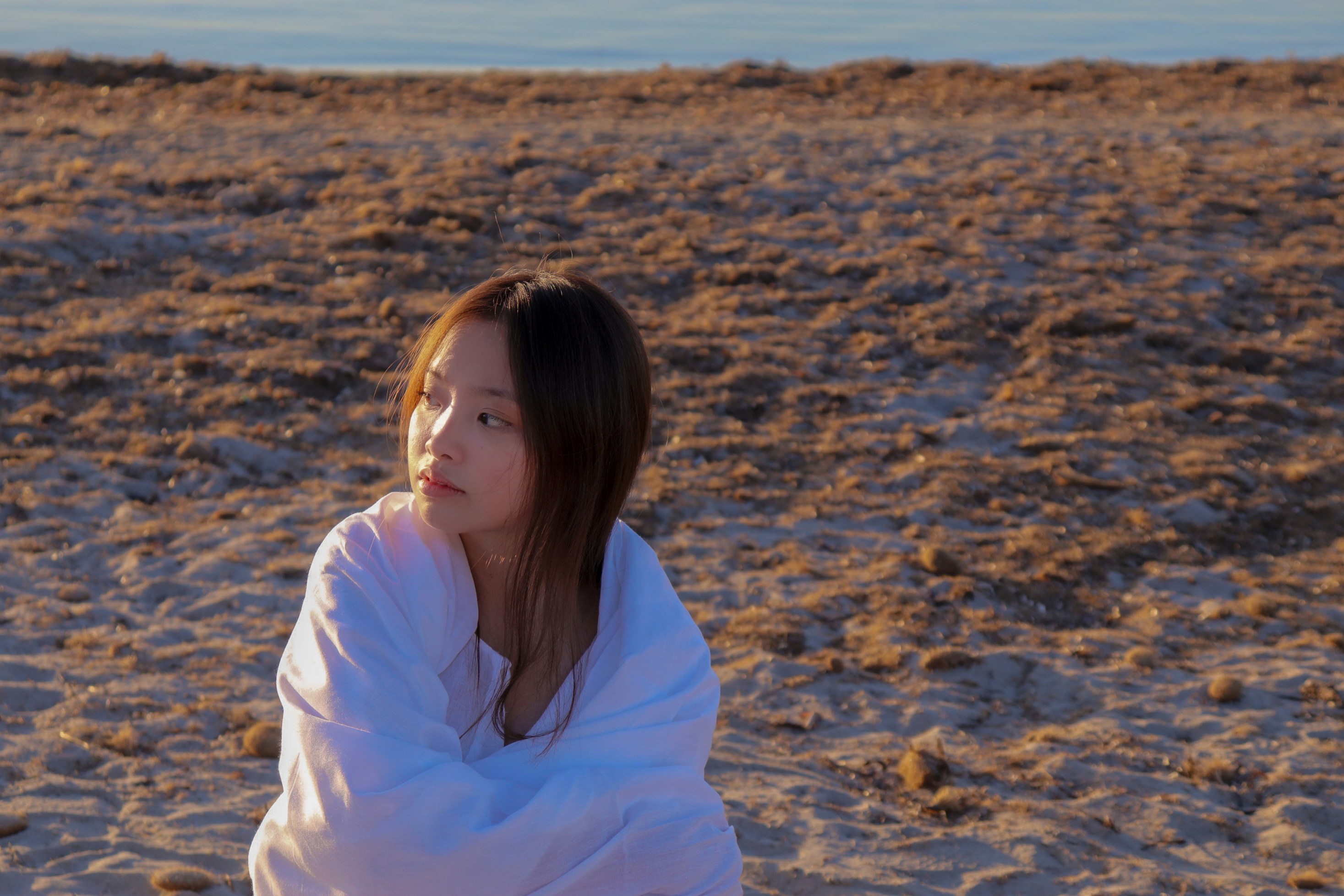 Person wrapped in a white blanket gazing thoughtfully on a sunlit beach.