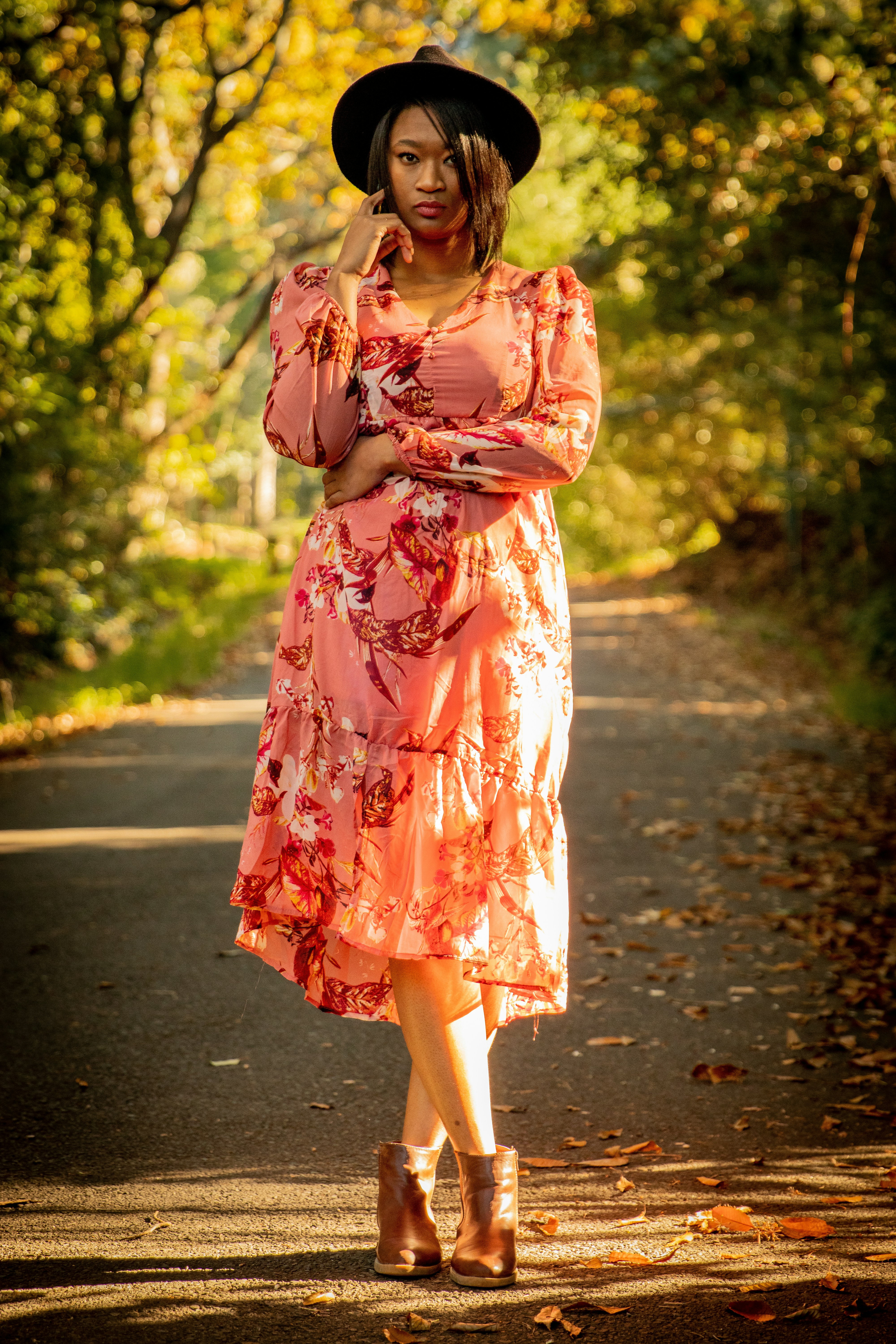 A woman in a floral dress stands confidently on a sunlit path, surrounded by autumn foliage. Her expression conveys strength and poise.