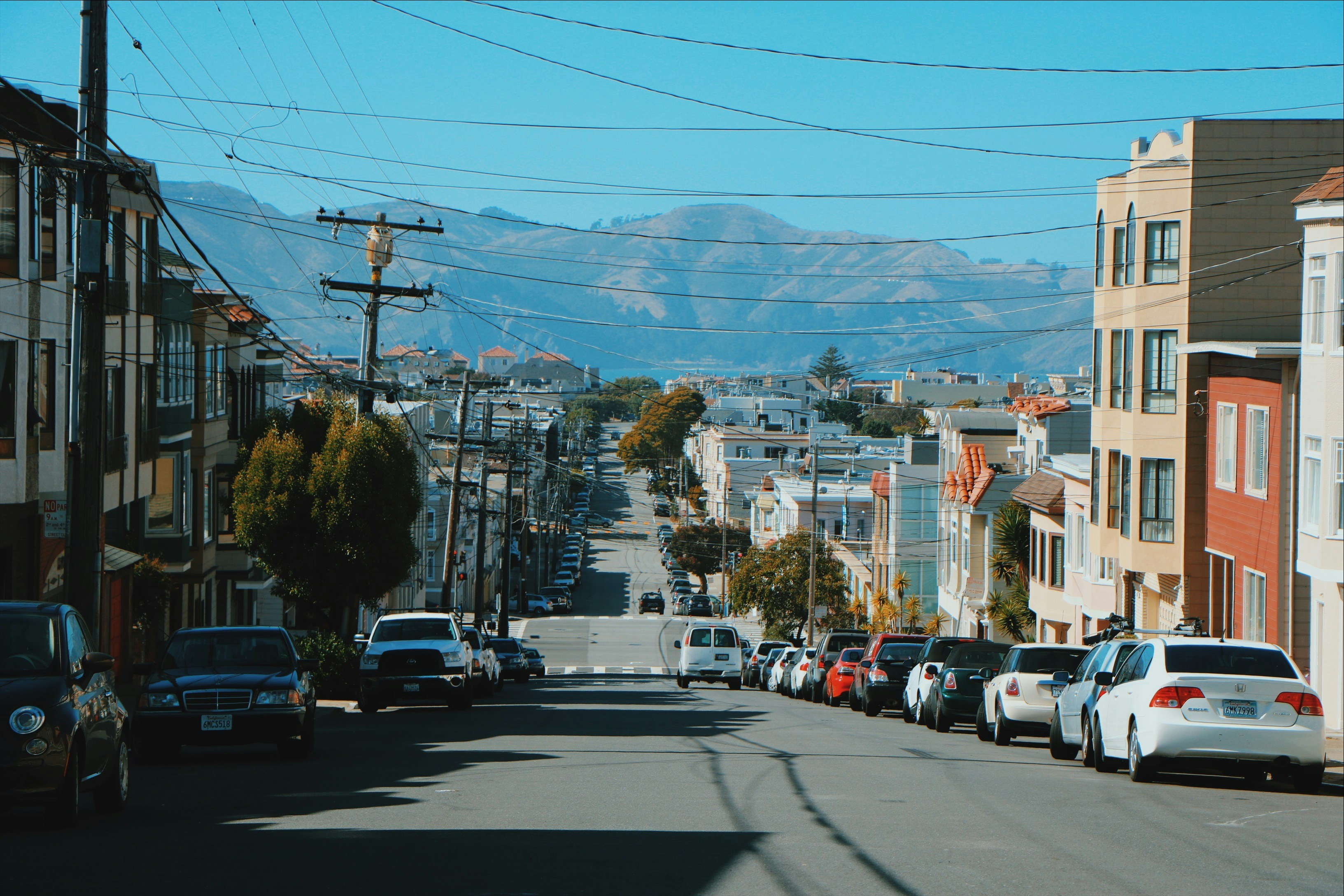Vibrant city street lined with parked cars, leading down towards distant mountains under a clear blue sky.