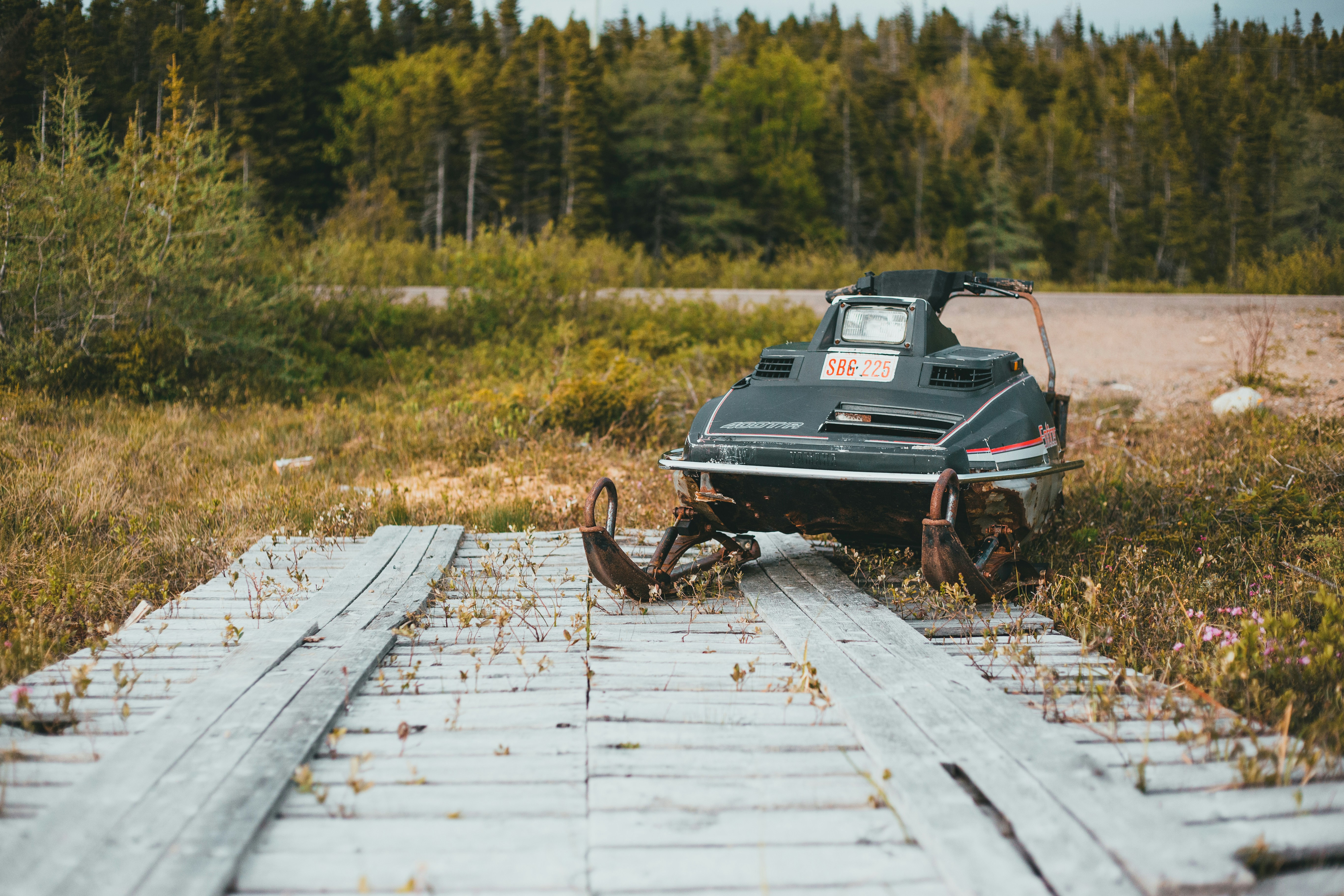 Abandoned snowmobile resting on a weathered wooden path, surrounded by lush greenery and distant trees. The scene evokes a sense of nostalgia and adventure.