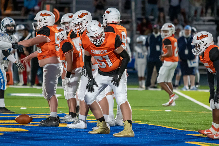 High school lineman #51 in orange and white crouched at the line on blue end-zone turf with teammates behind him.