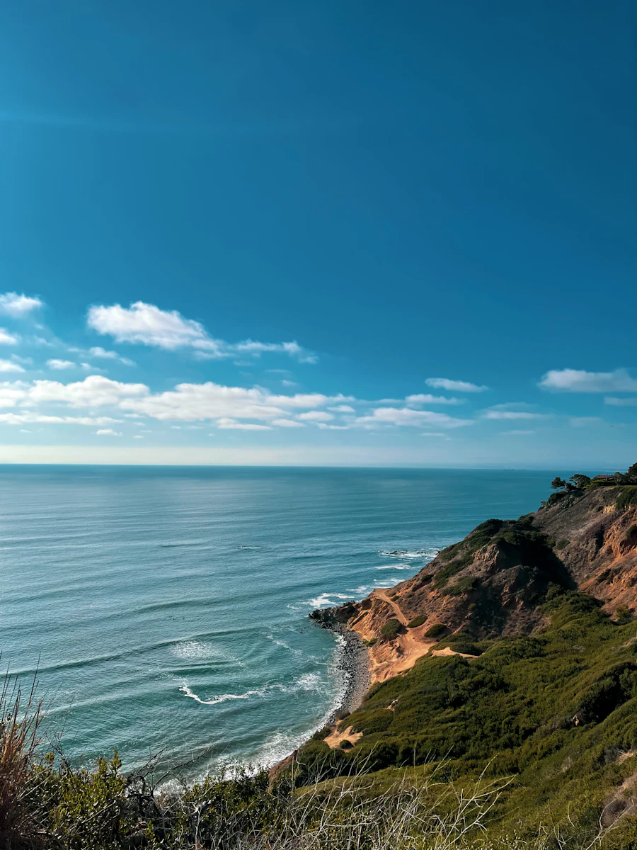 Sweeping coastal cliff with winding dirt path above a calm ocean and scattered clouds. The shoreline curves around the headland under a bright blue sky.