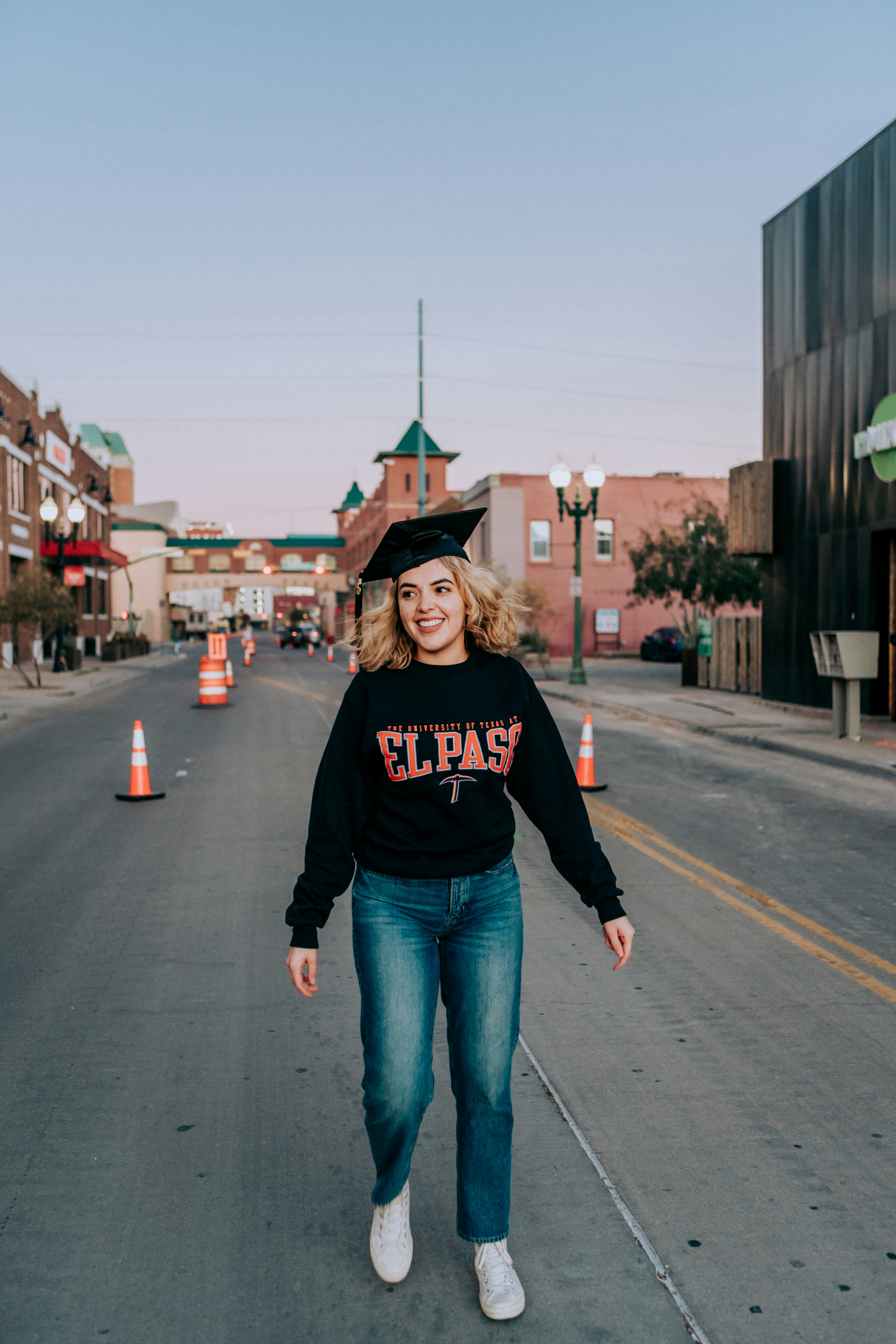 Graduate joyfully walking down a deserted street in El Paso, wearing a cap and gown, with festive decorations in the background.
