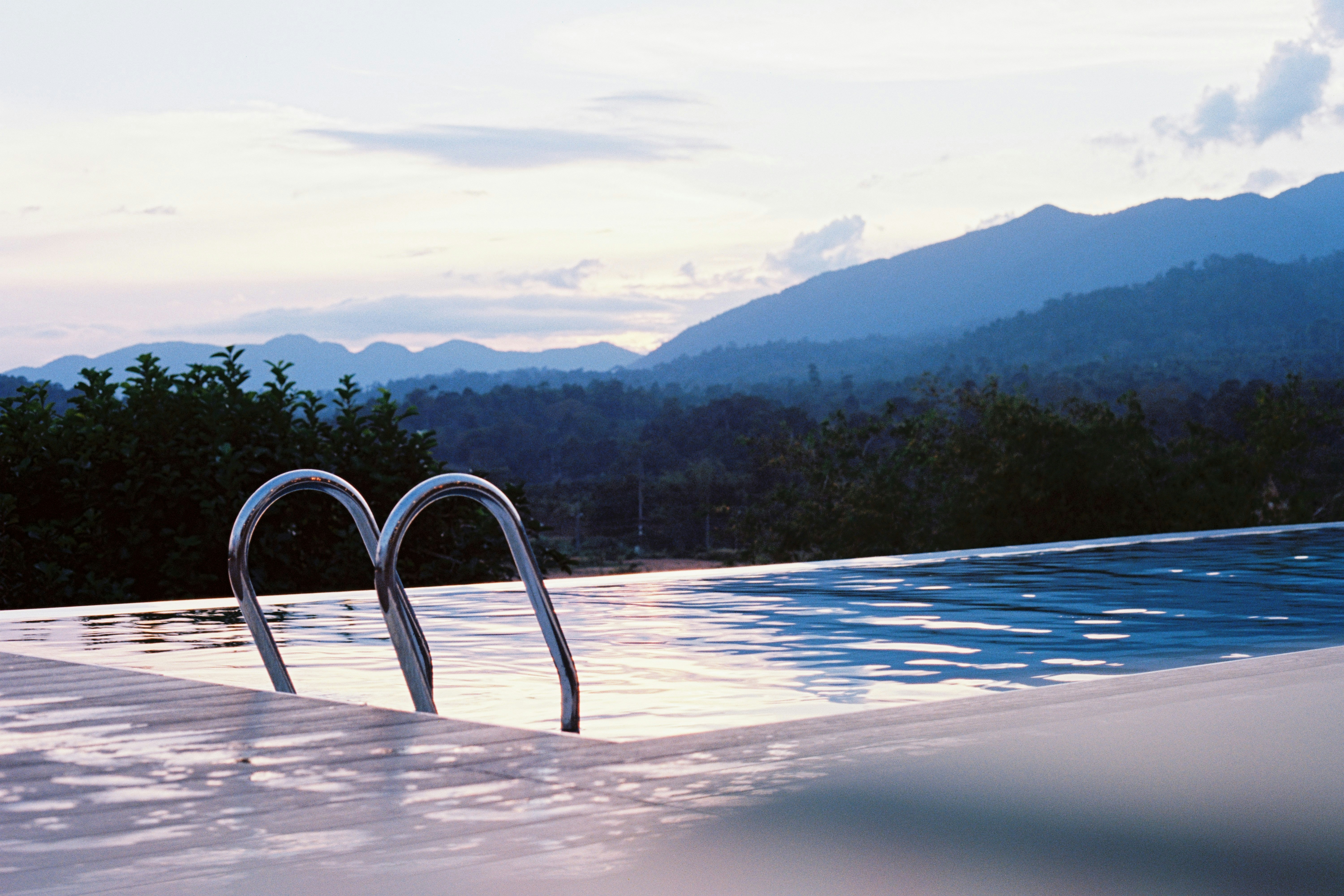 Photograph of an infinity-edge pool at dusk, with metal ladder arches in the foreground and a distant mountain range.
