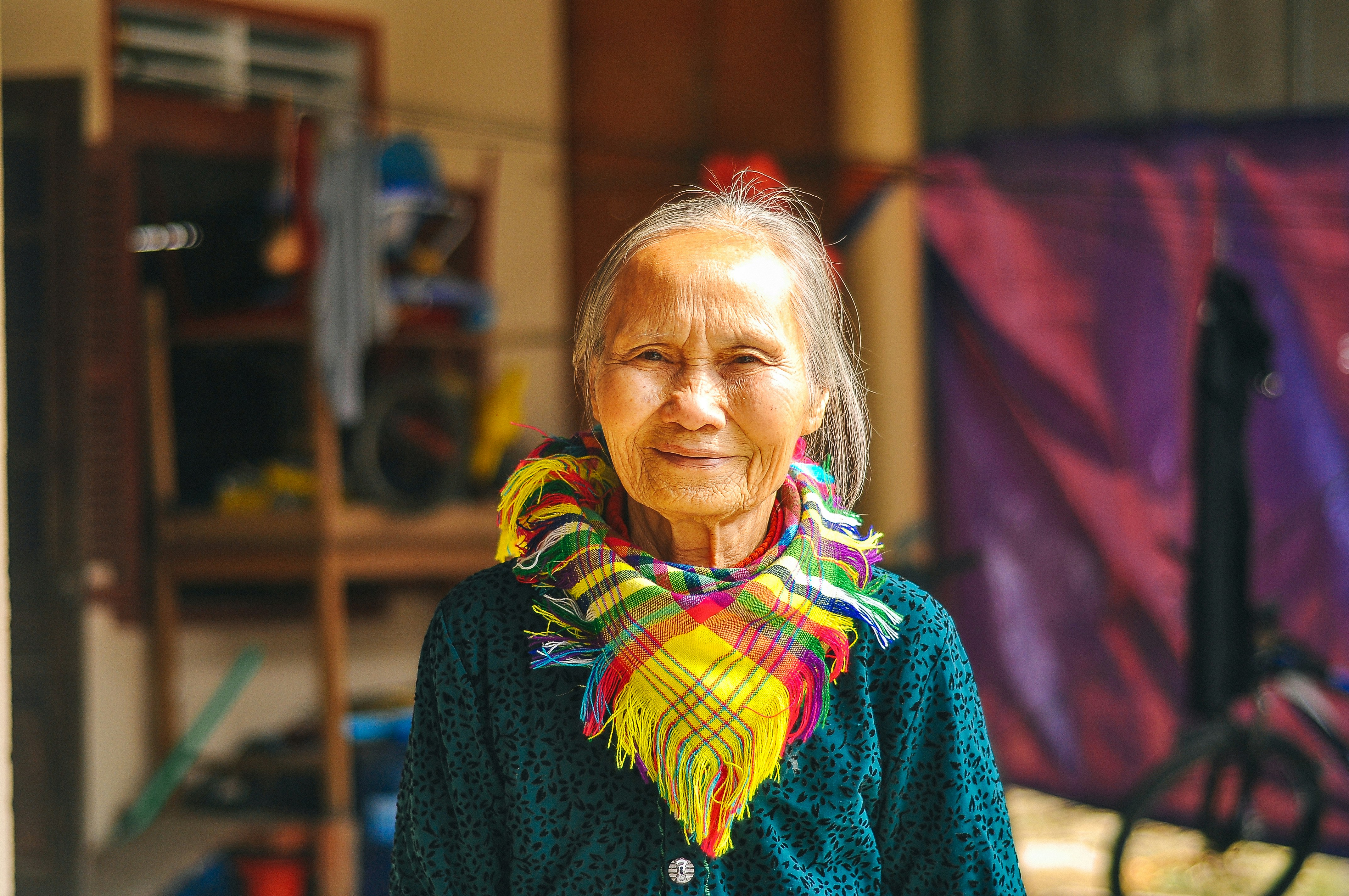 Elderly woman with a vibrant scarf smiling warmly, set against a rustic background with hints of everyday life.