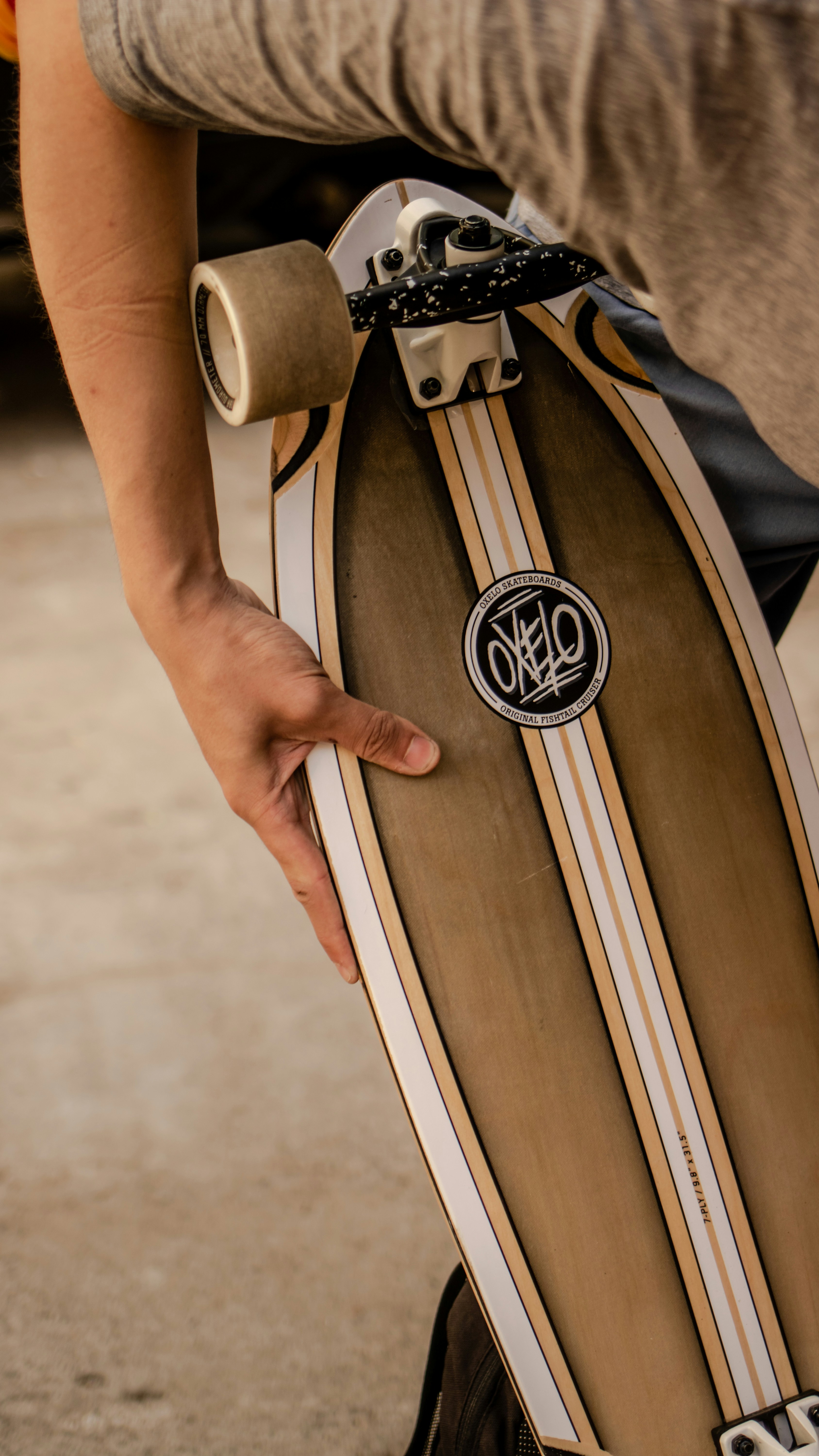 A hand gripping a stylish longboard, showcasing its intricate design and wheels against a textured surface. The focus is on the rider's anticipation before the ride.