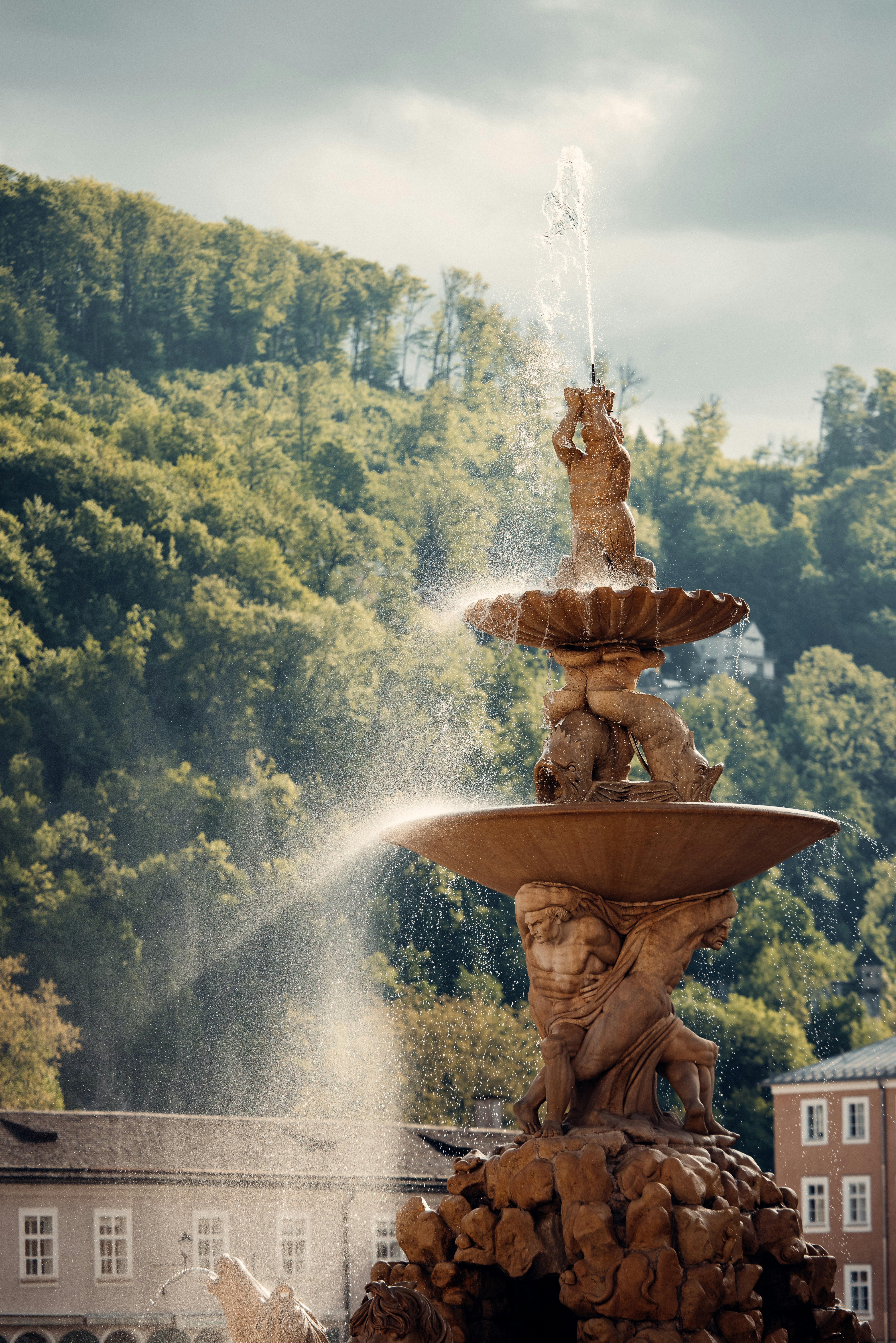 Intricate stone fountain with cascading water, set against a backdrop of lush green hills. The scene captures the essence of tranquility and artistry.