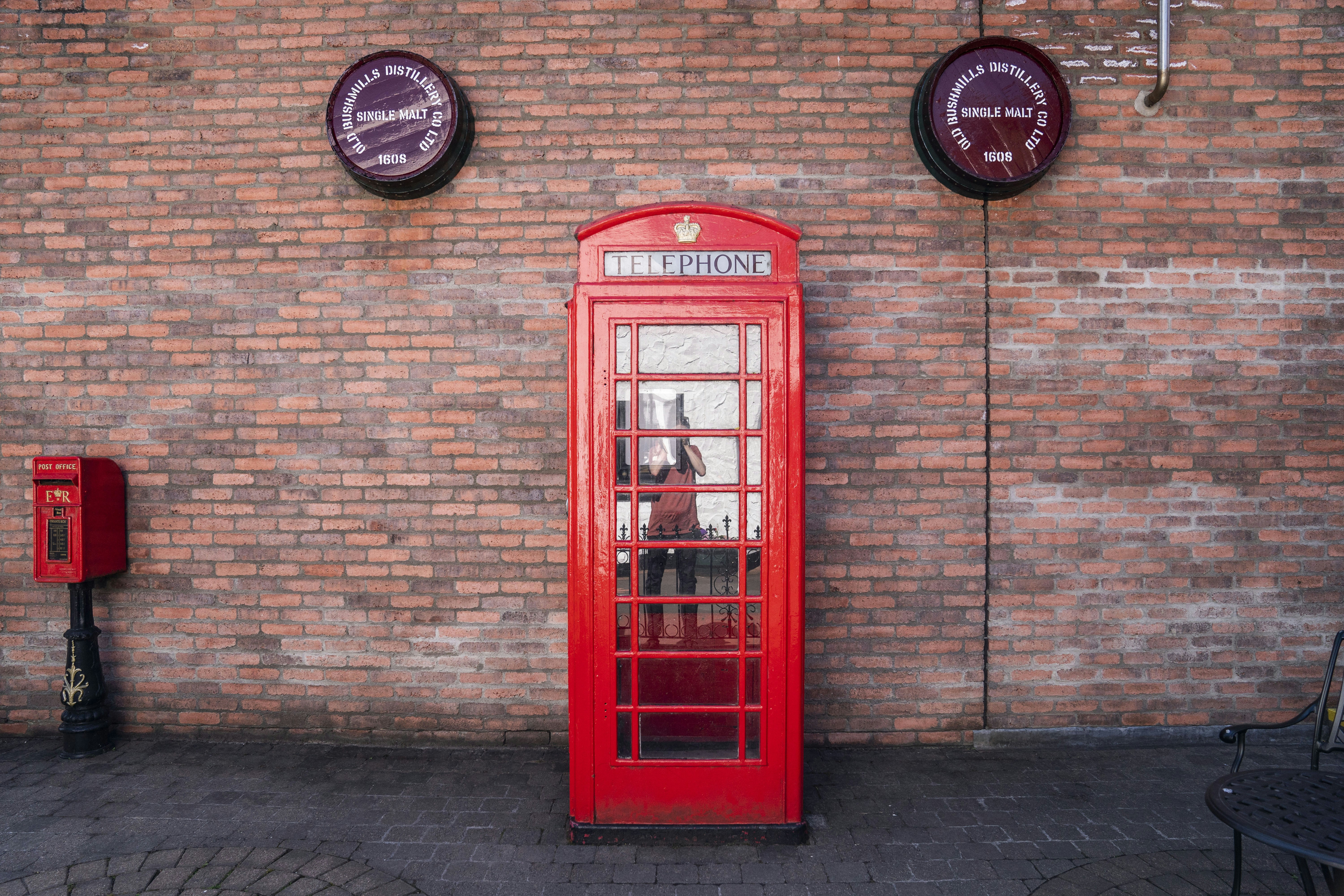 A classic red telephone booth stands against a textured brick wall, flanked by vintage circular signs and a post box, evoking nostalgia for a simpler time.