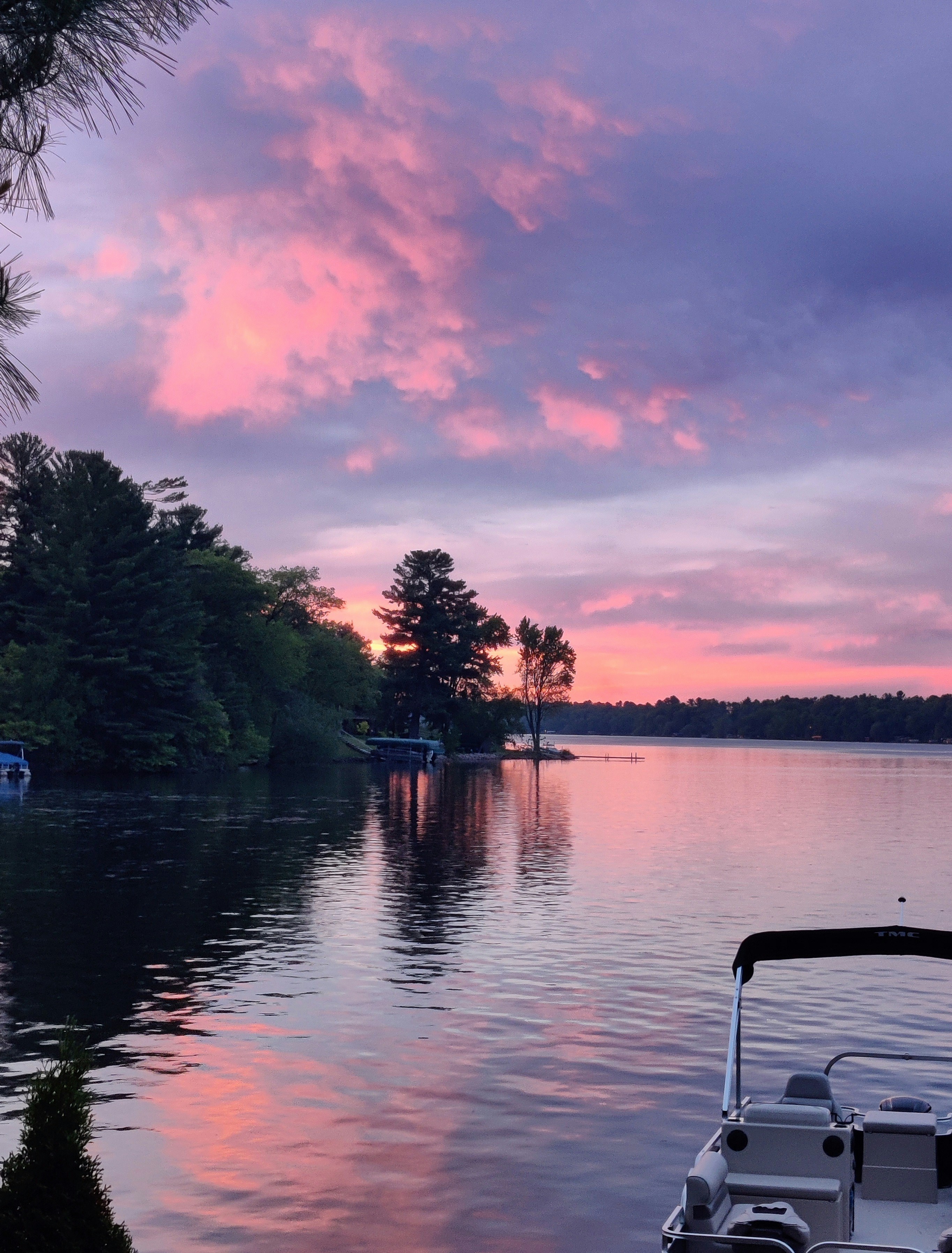 Vibrant sunset colors reflecting on a tranquil lake, framed by lush trees and a boat at the shore.