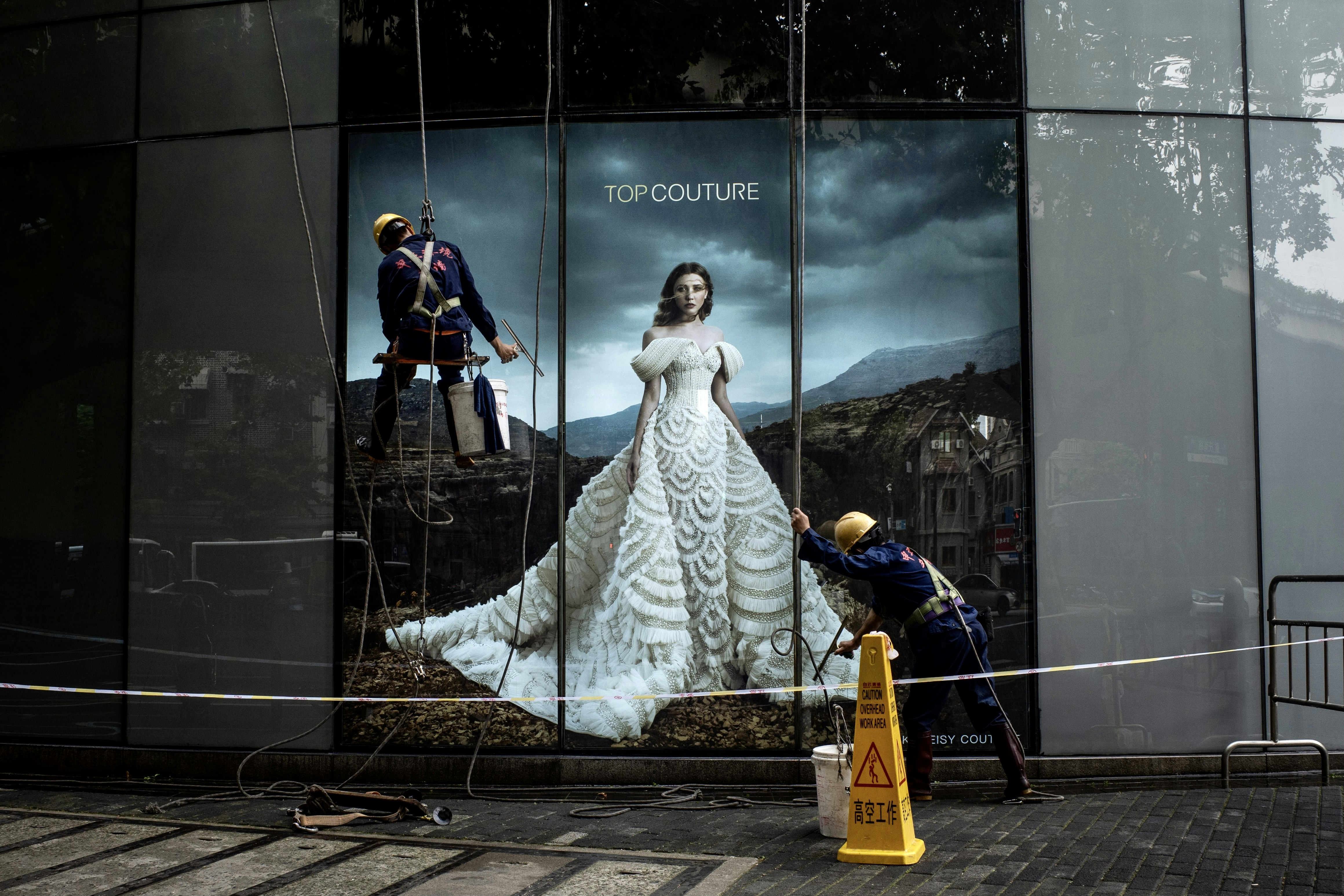 Workers clean a large fashion advertisement on a city building, showcasing a woman in a white gown against a dramatic sky.