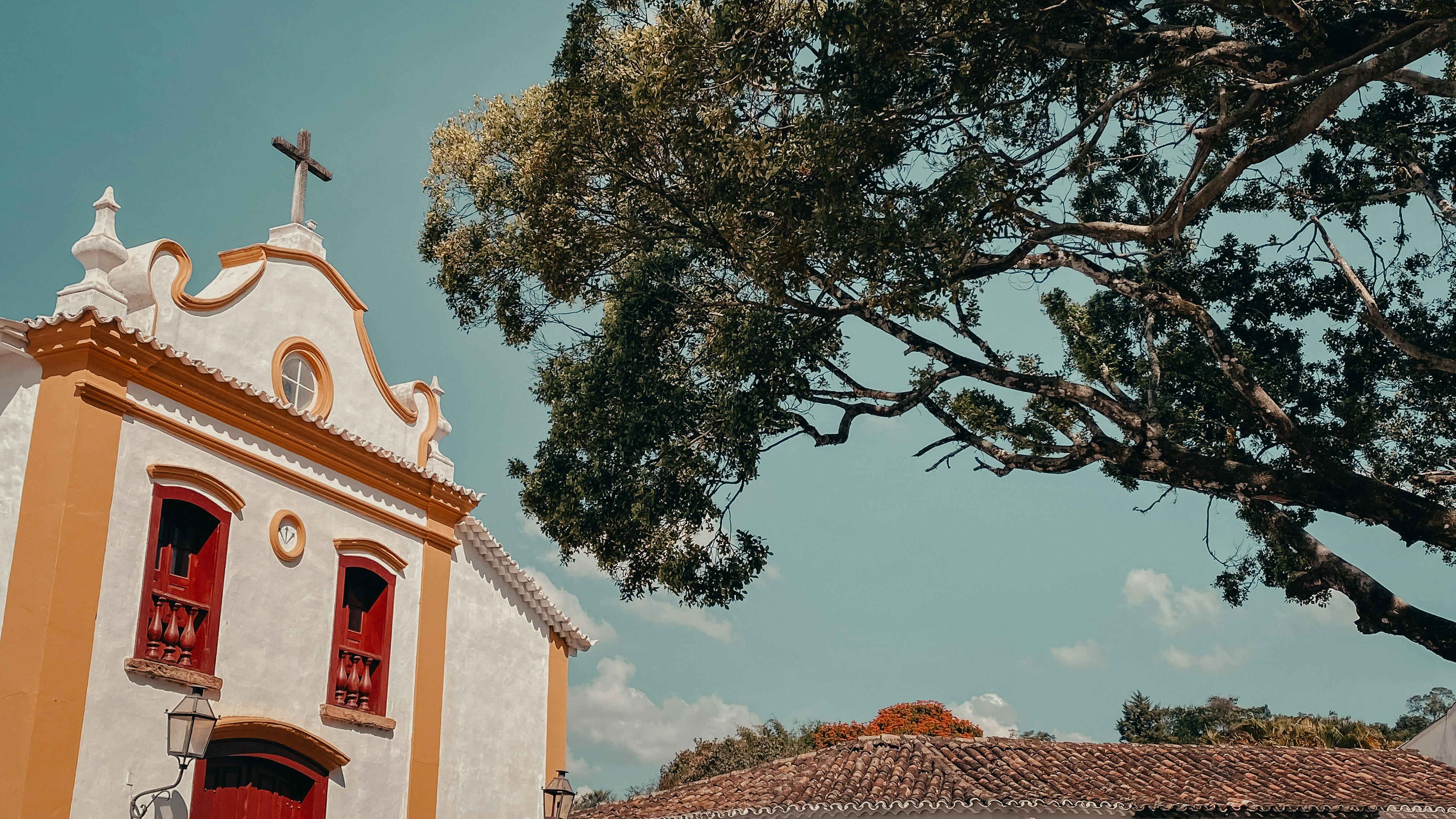 Historic church with vibrant red accents under a clear blue sky, framed by lush greenery and a sprawling tree. The architecture reflects a rich cultural heritage.