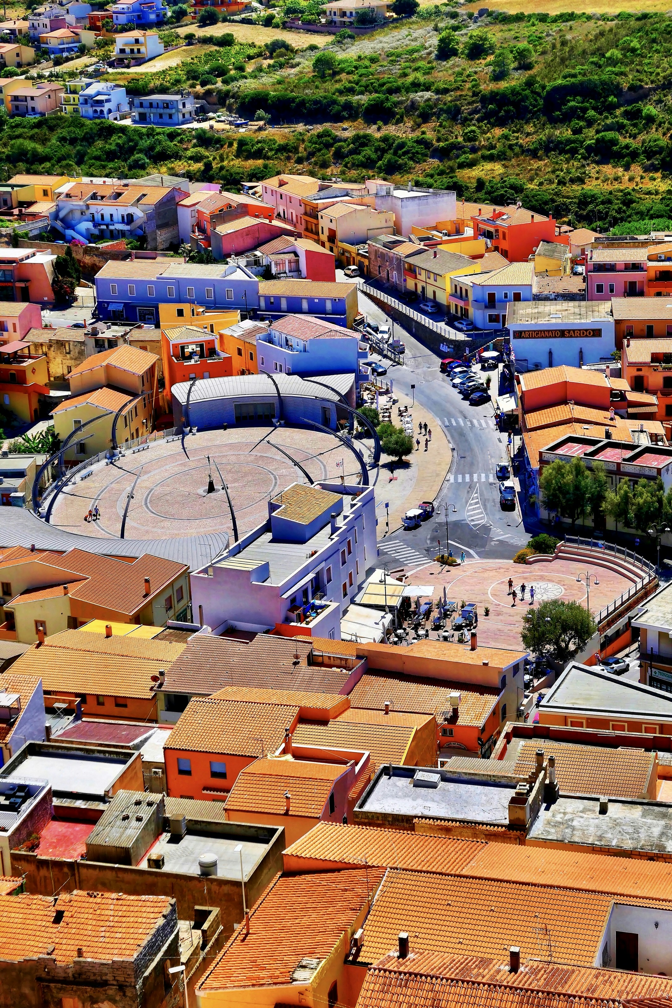Vibrant rooftops and streets of a coastal town captured from above, showcasing a lively square and colorful architecture. 