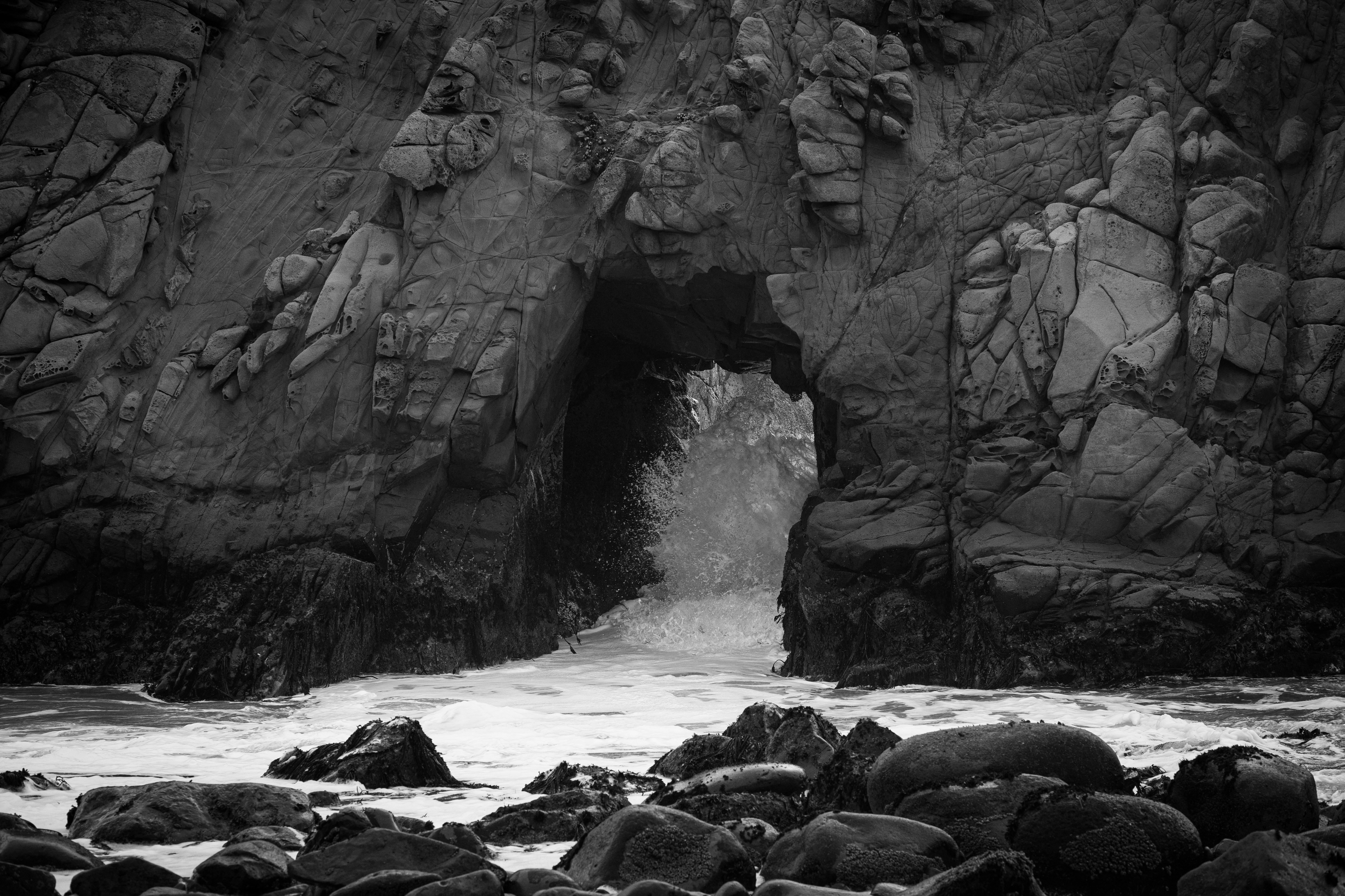Rocky archway framing crashing waves, with textured cliffs and scattered boulders in the foreground.