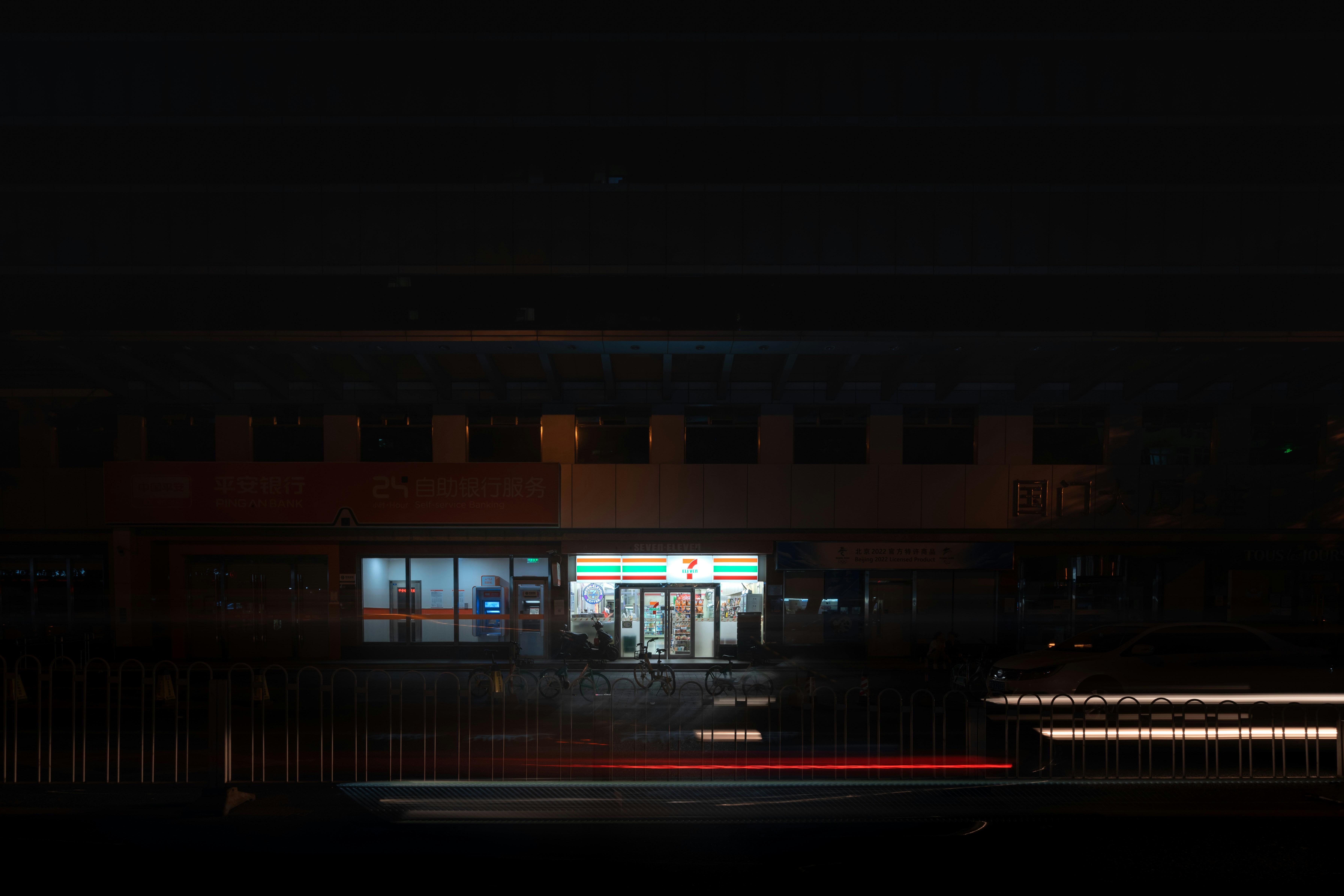 A convenience store illuminated against a dark urban backdrop, showcasing the contrast between light and shadow. The scene captures the essence of city life at night.