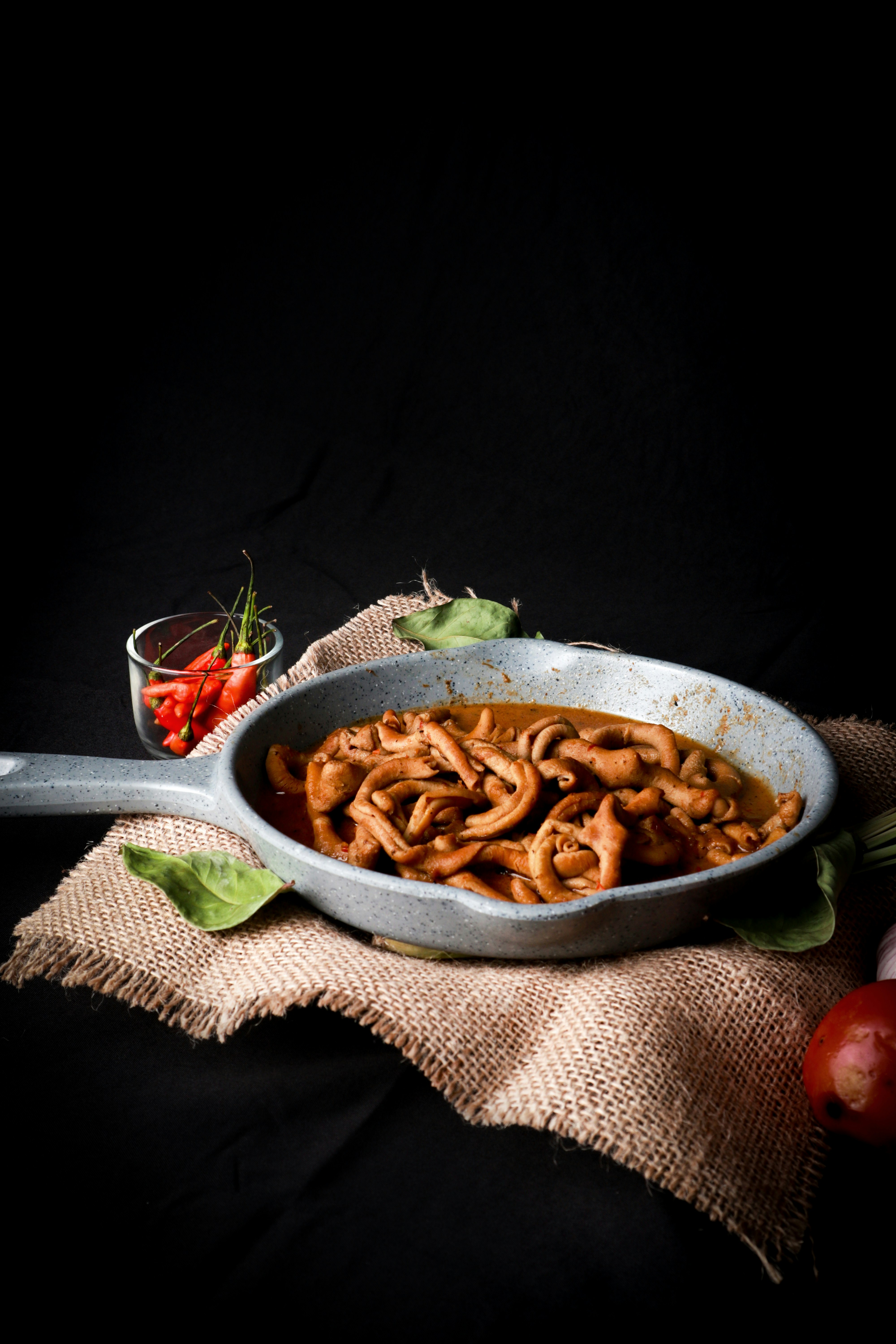 Mushrooms simmer in a gray skillet set on a burlap cloth with a small glass of peppers in the background; rustic dinner scene with a dark backdrop.
