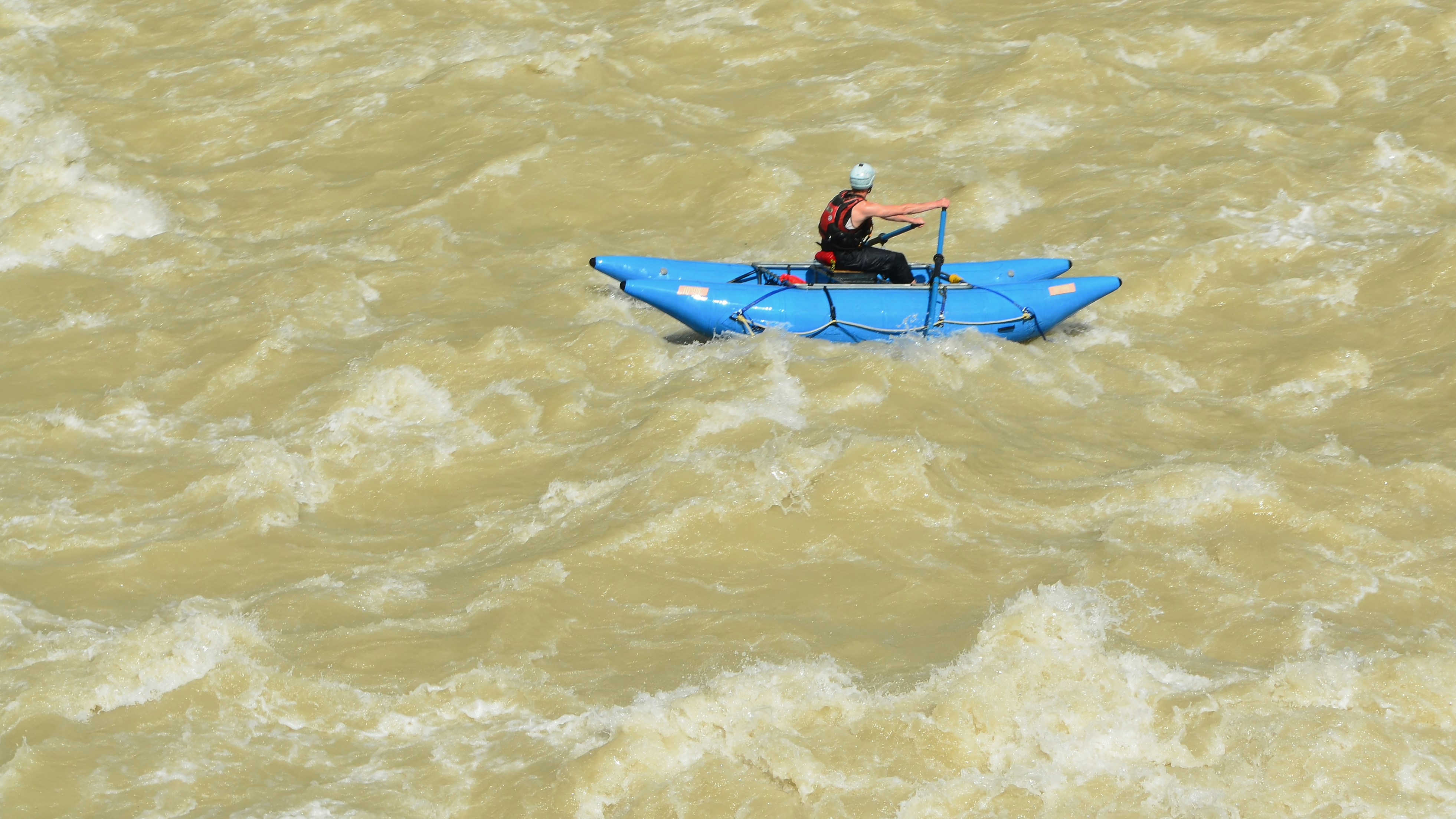 A lone rafter expertly maneuvers a bright blue raft through swirling, muddy river rapids. The scene captures the thrill of adventure and the power of nature.