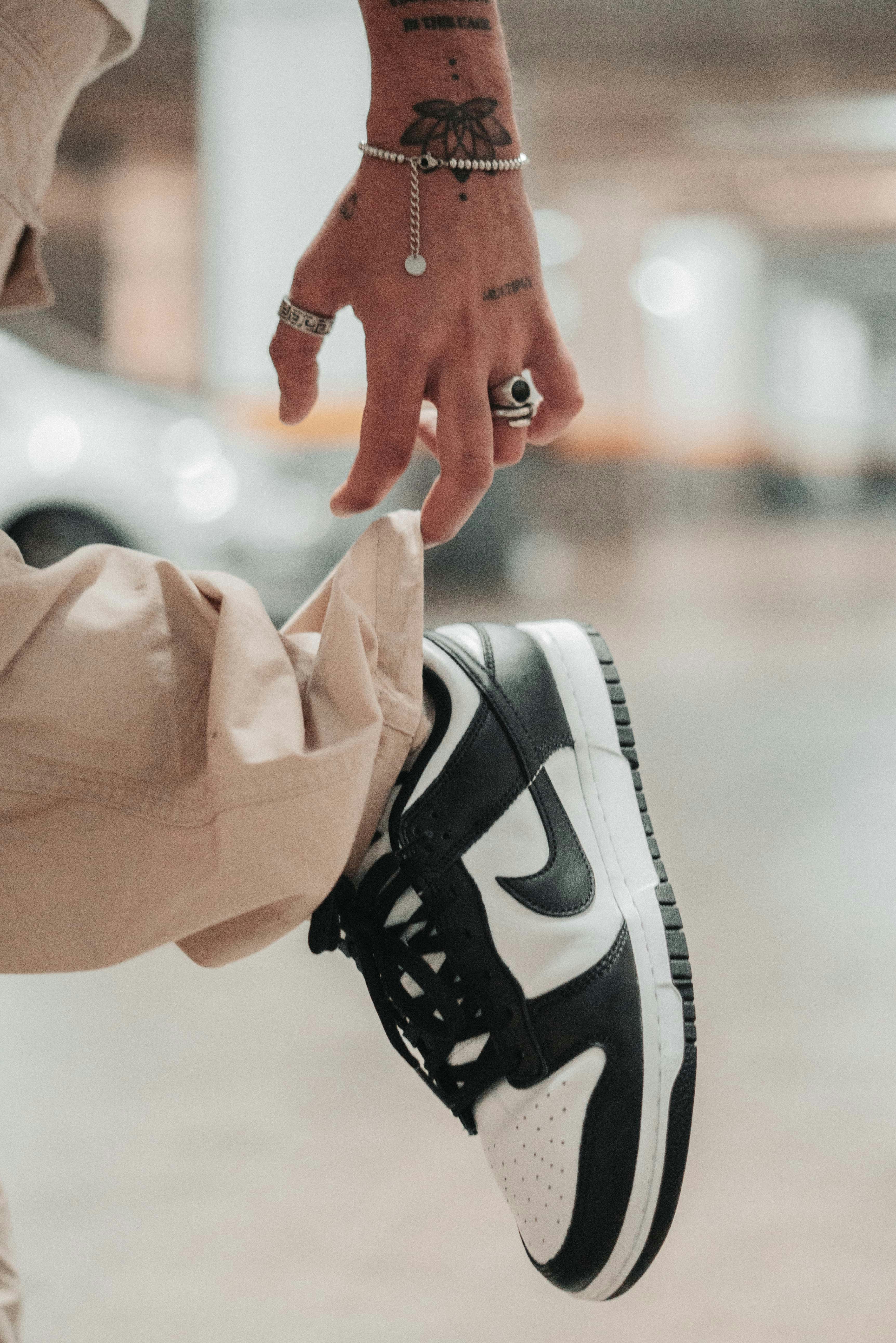 Close-up of a person’s foot wearing a black and white sneaker, with a hand adorned with rings and bracelets extending into the frame. The background features a blurred parking garage setting.