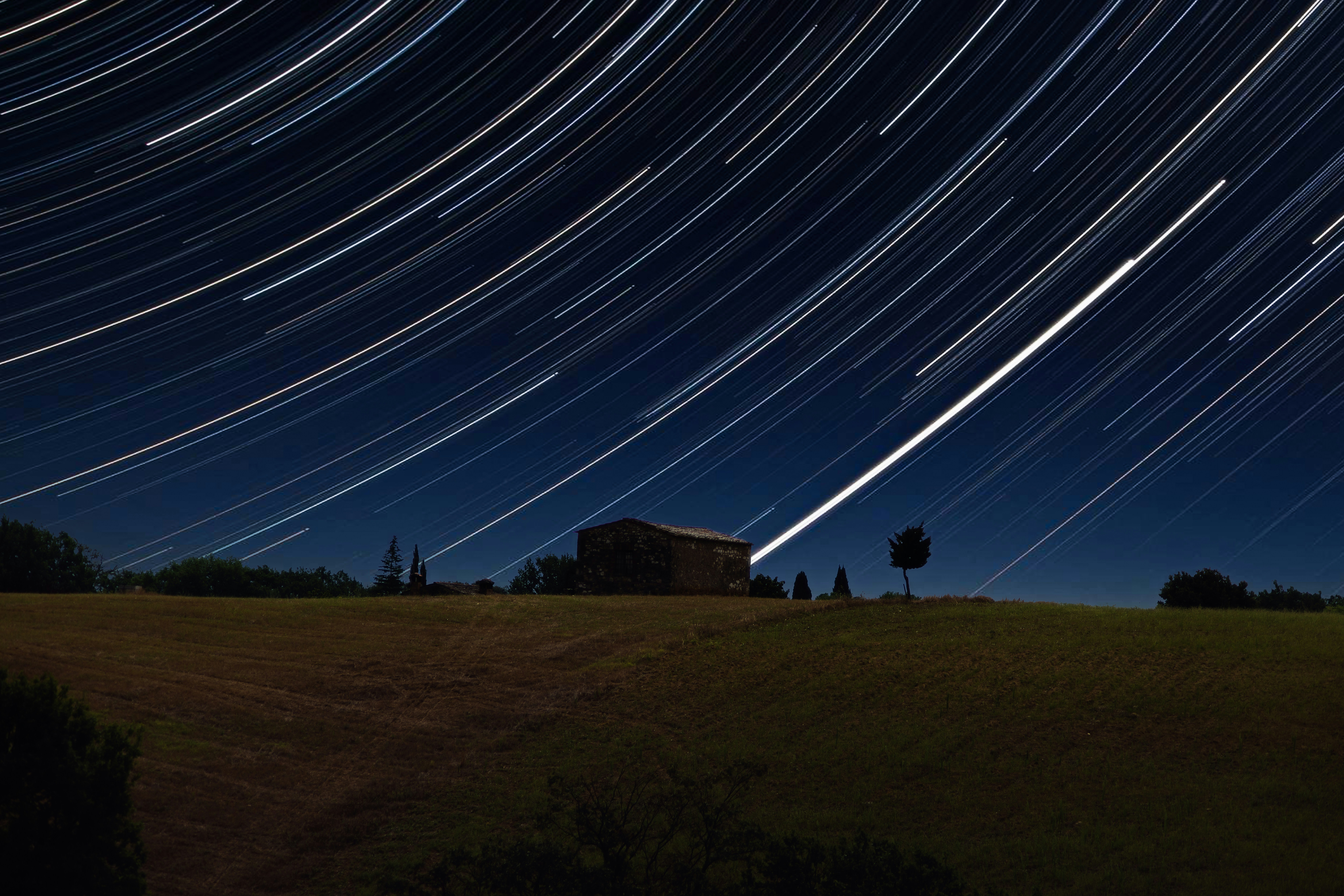 Star trails arc across a clear night sky above a solitary house on a grassy hill.