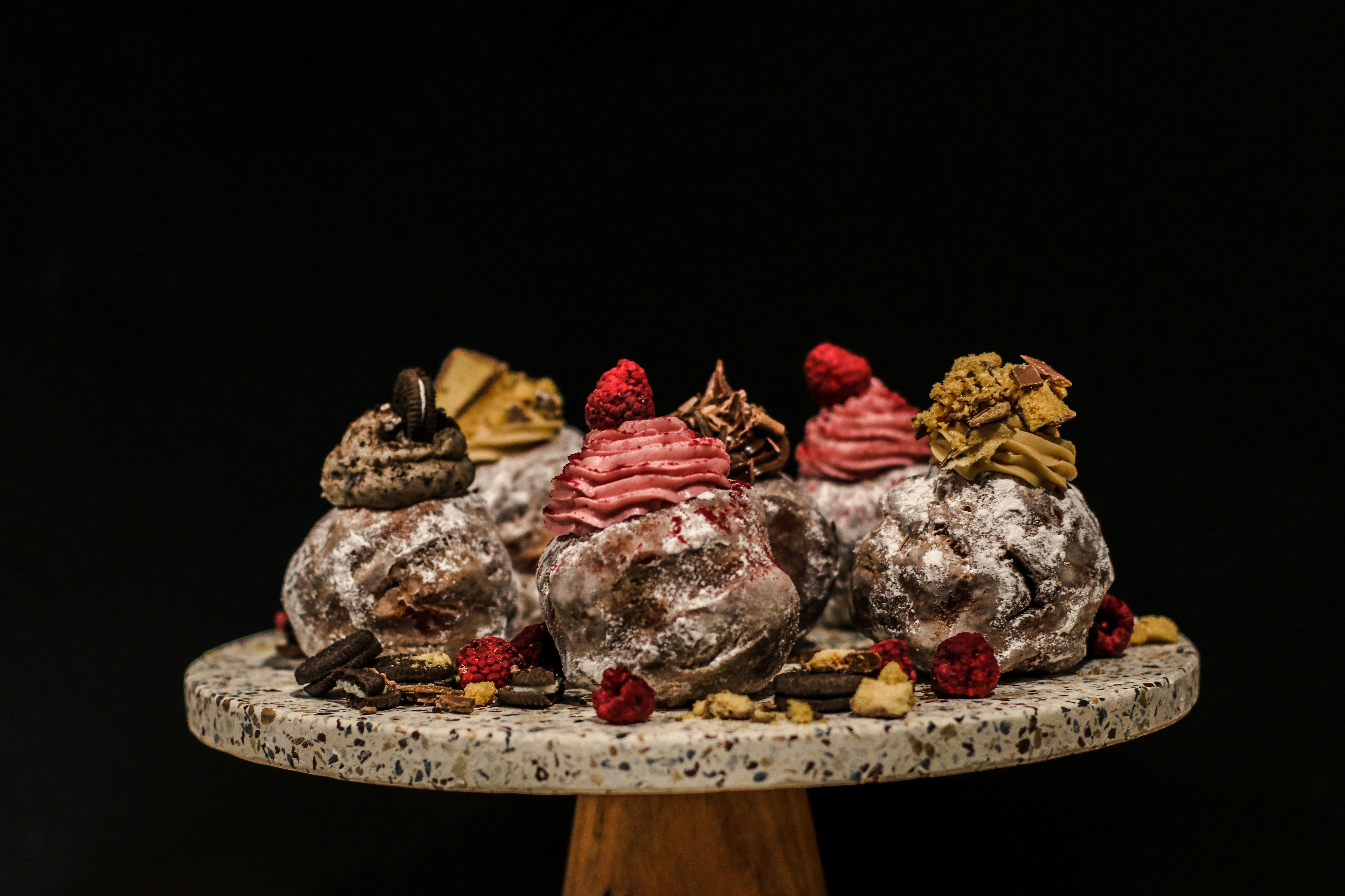 A beautifully arranged display of powdered pastries topped with colorful frosting and garnishes on a textured cake stand.