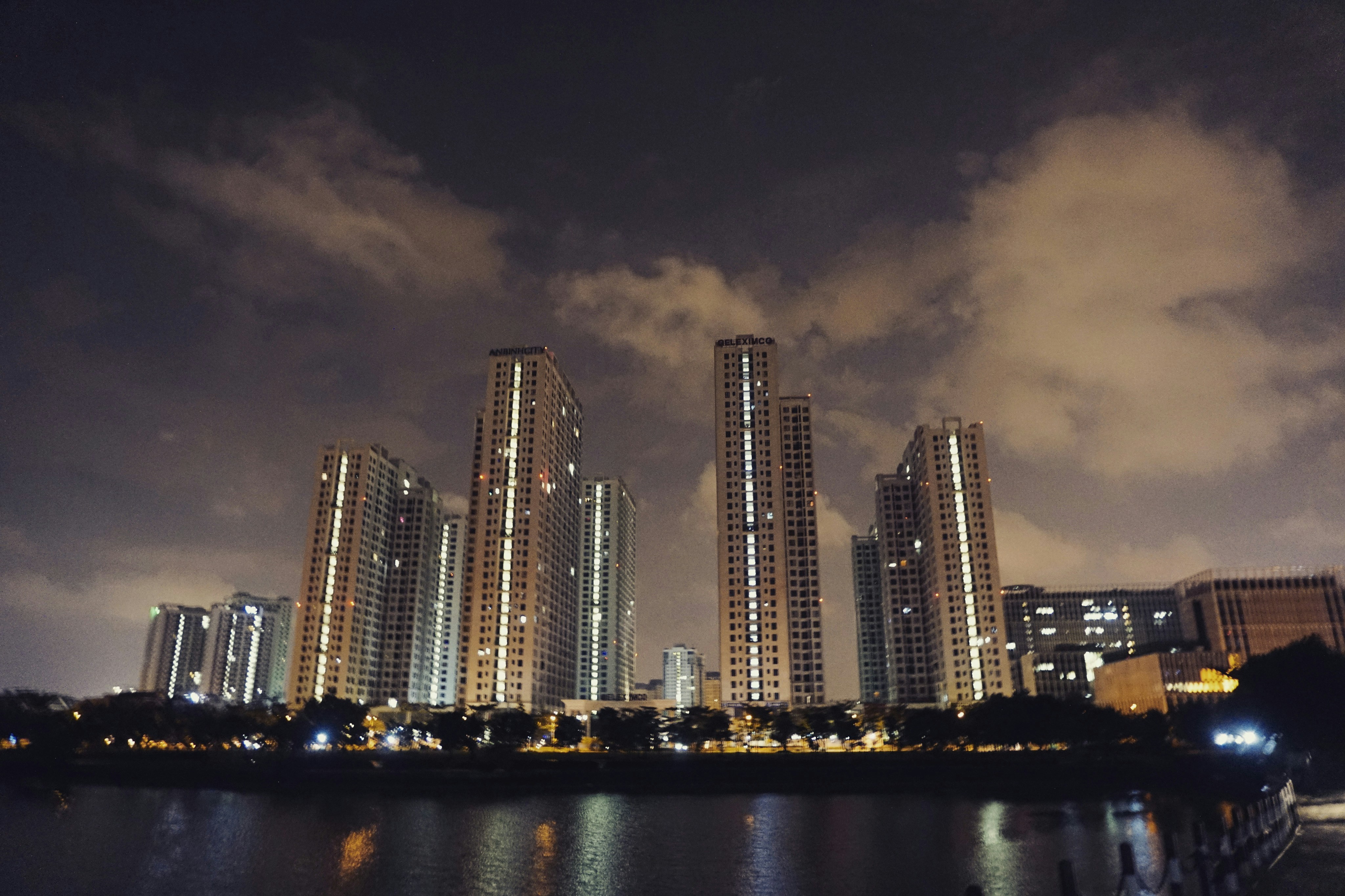 Illuminated skyscrapers reflect on a calm river under a cloudy night sky.