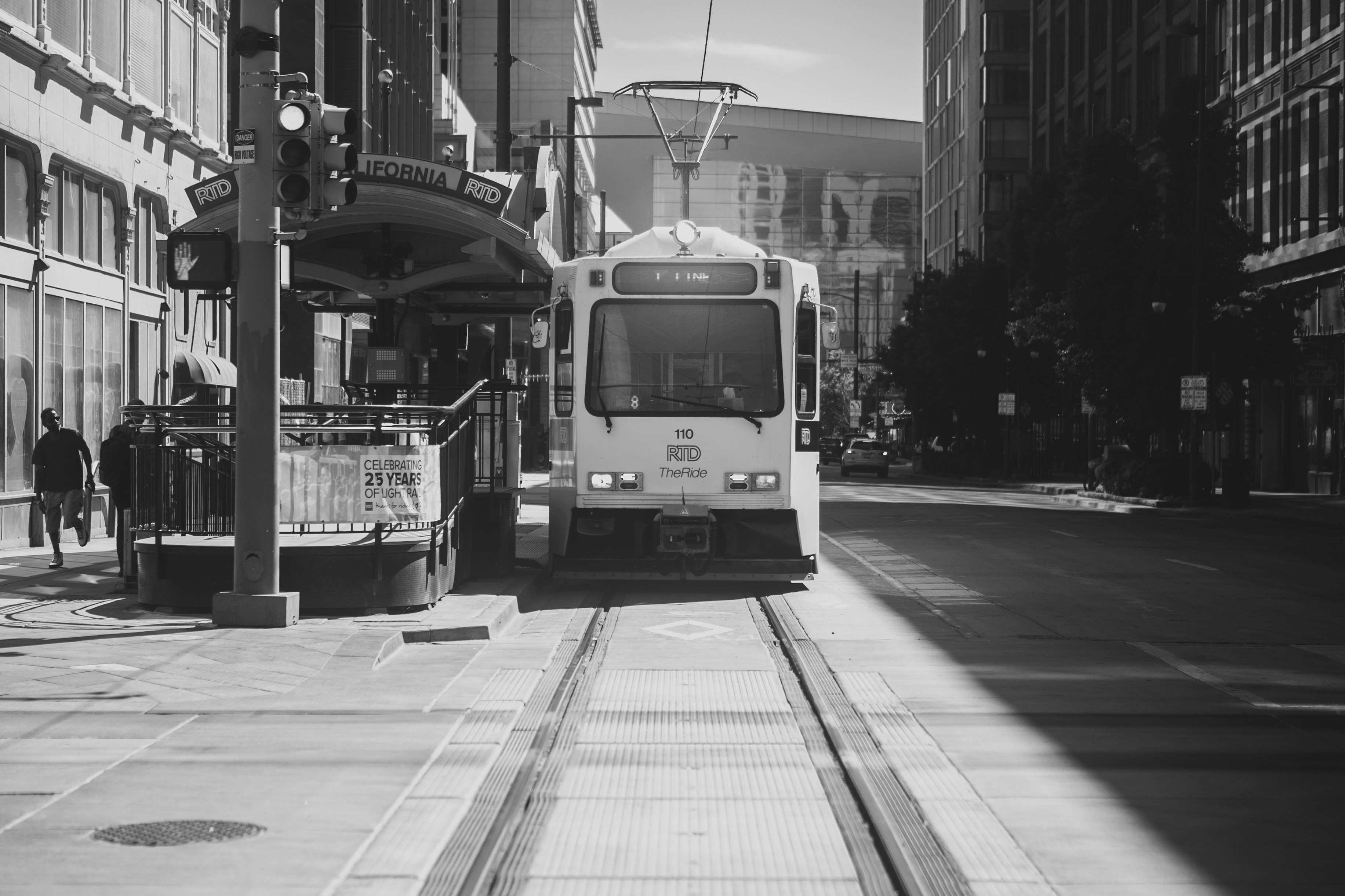 Light rail train approaching a transit stop in a bustling urban environment, framed by modern architecture and shadows.