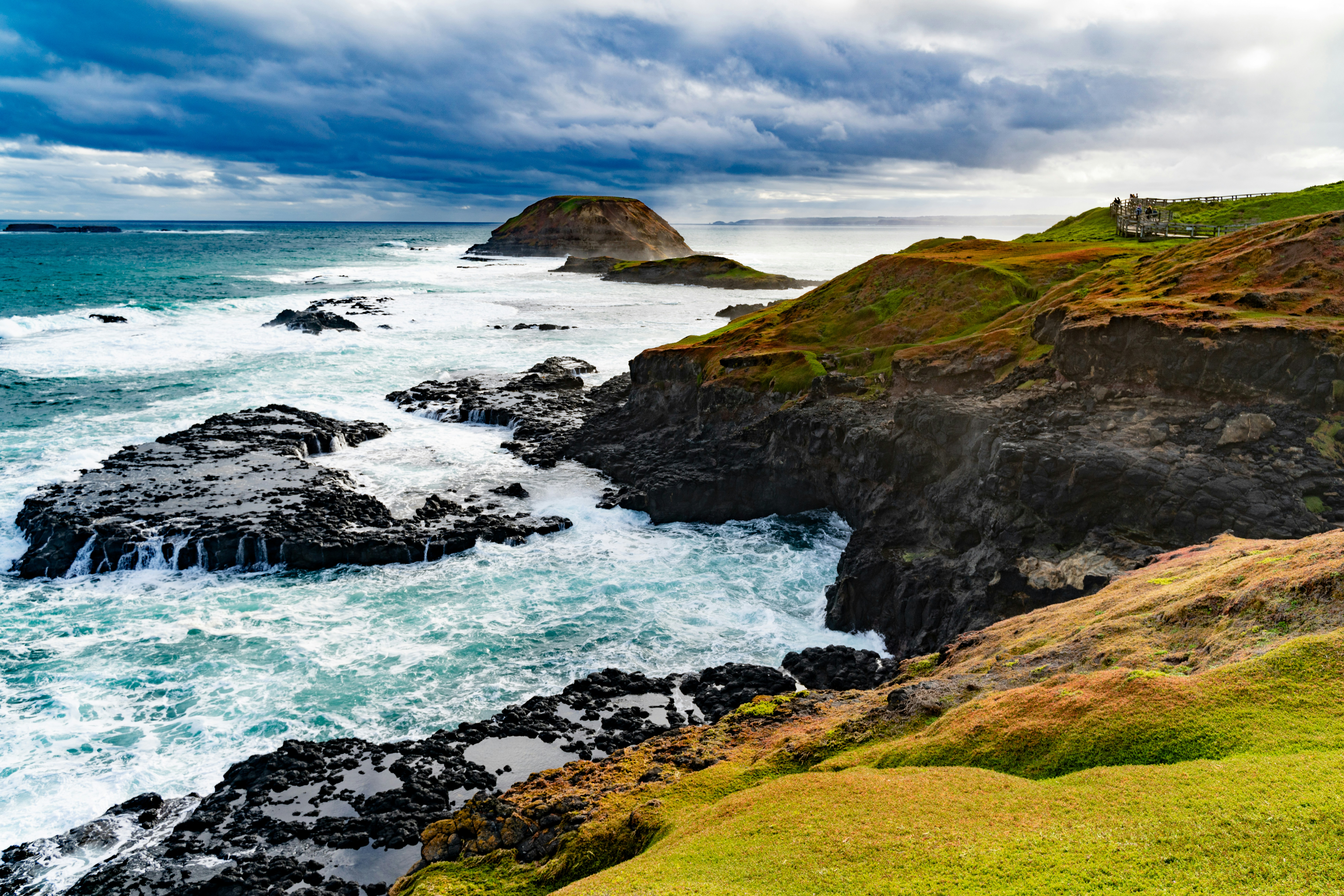Dramatic rocky coastline with waves crashing against the cliffs and a solitary house perched atop the grassy land.