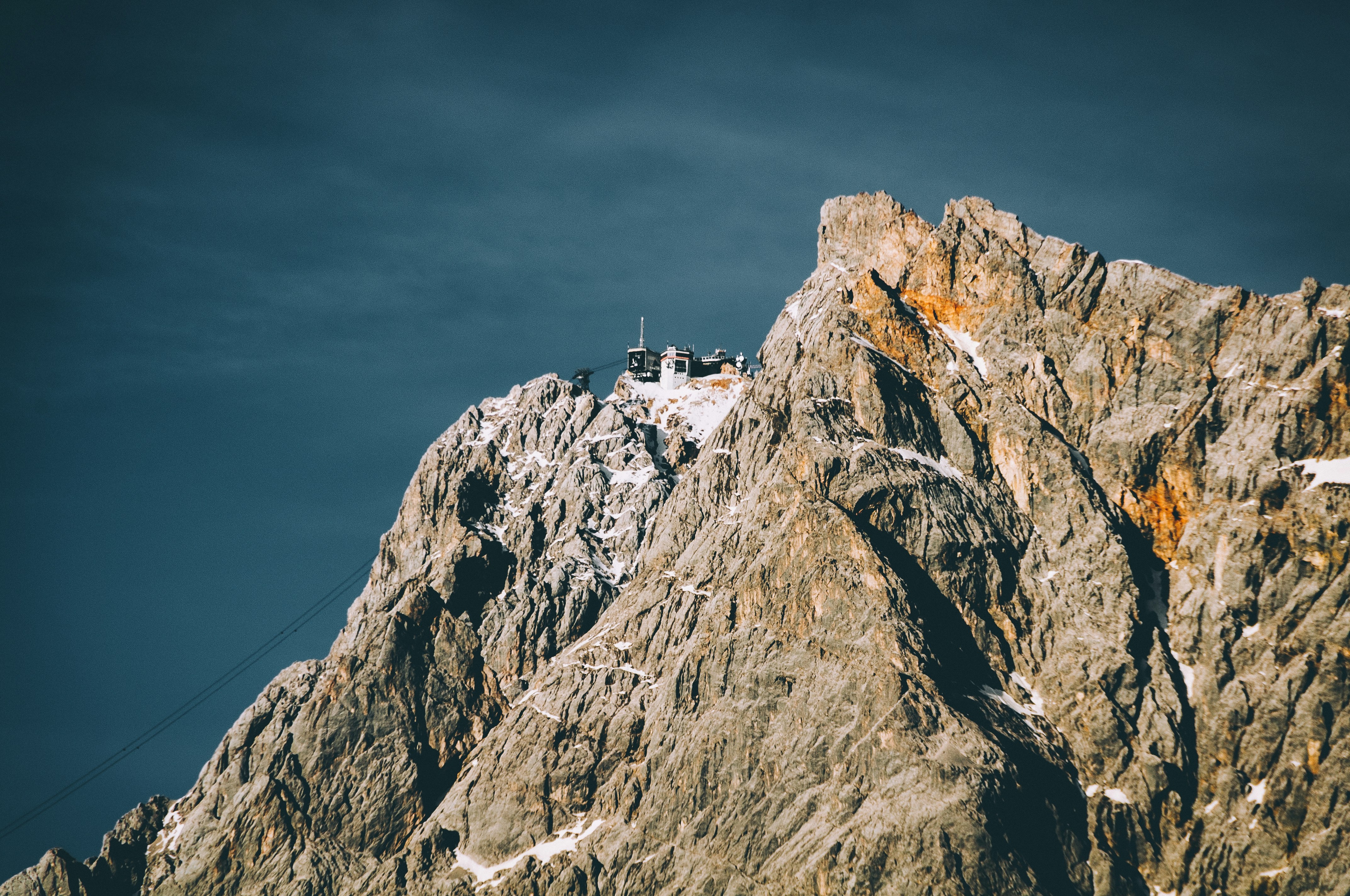 Rugged mountain summit with a small outpost and antenna rises against a steel-blue sky. The scene highlights the contrast between jagged rock and human-made structures.