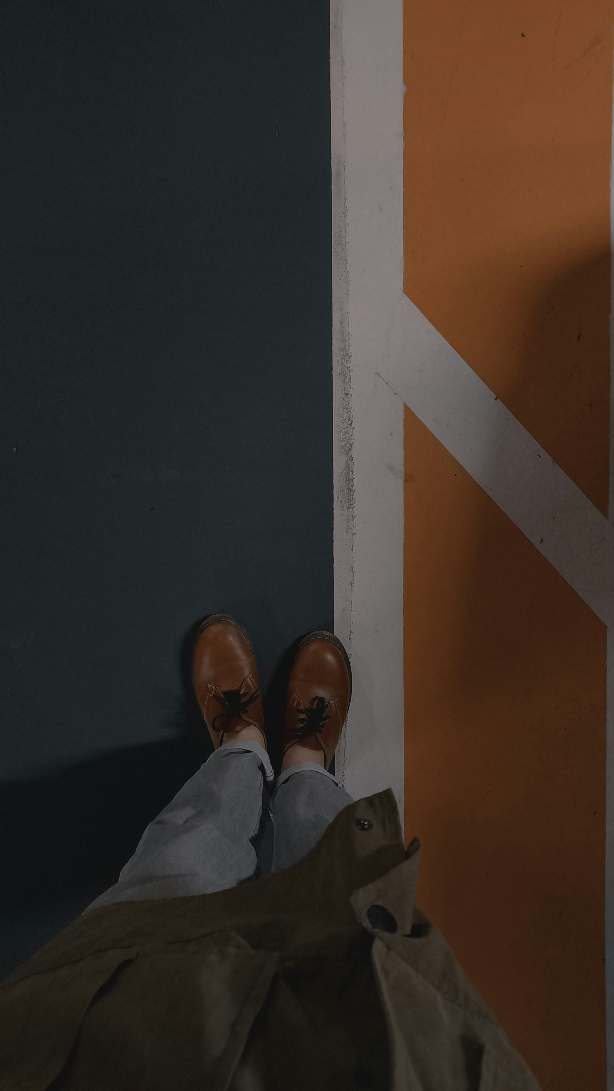 A pair of brown shoes stands on a contrasting blue and orange floor, framed by a stylish trench coat. The composition emphasizes the interplay of colors and textures.