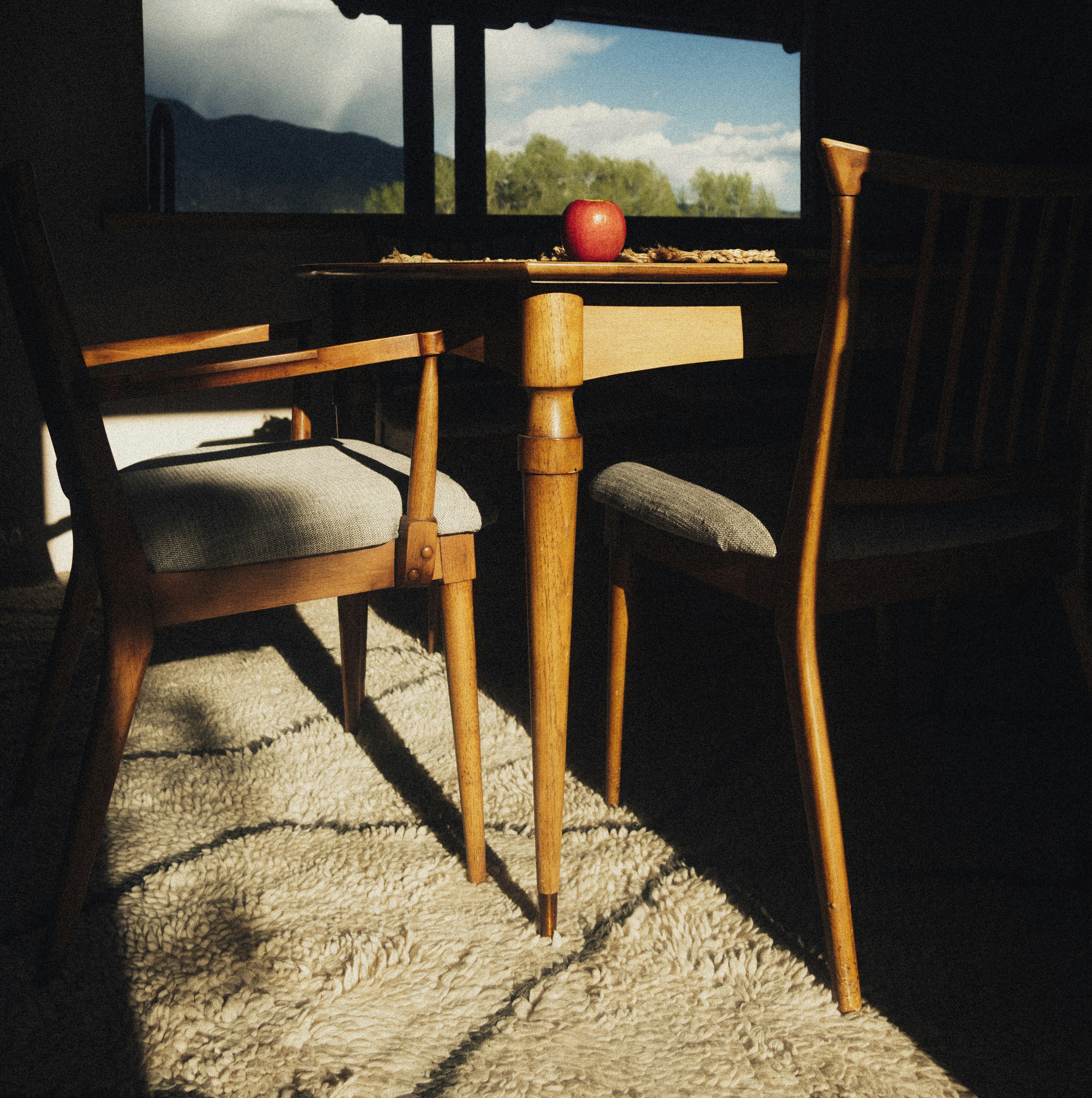 A wooden table set with an apple and surrounded by two chairs, illuminated by soft sunlight streaming through a window with a view of mountains.