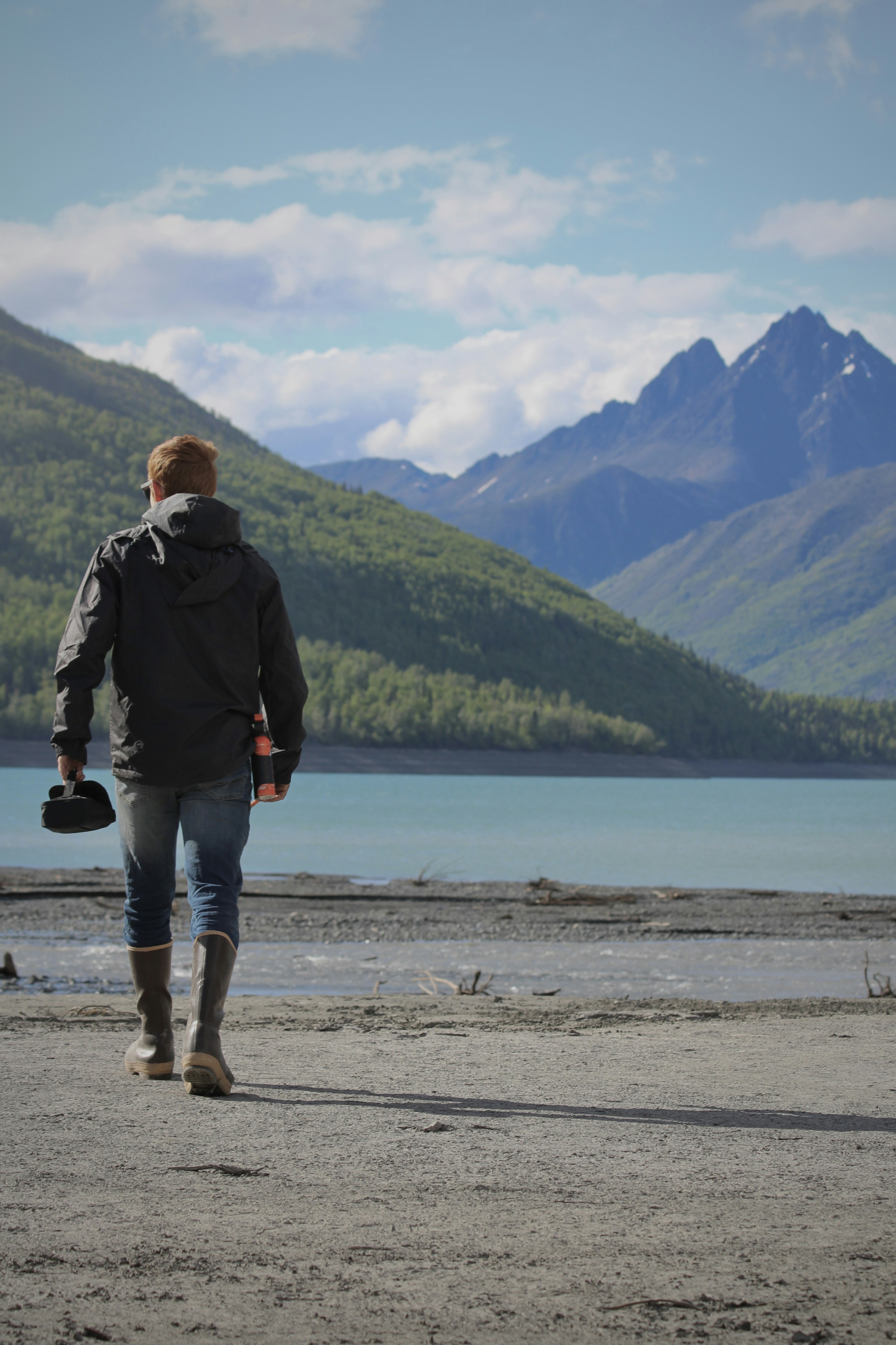 A figure walks along a sandy shore, holding a camera, with majestic mountains and a tranquil lake in the background.