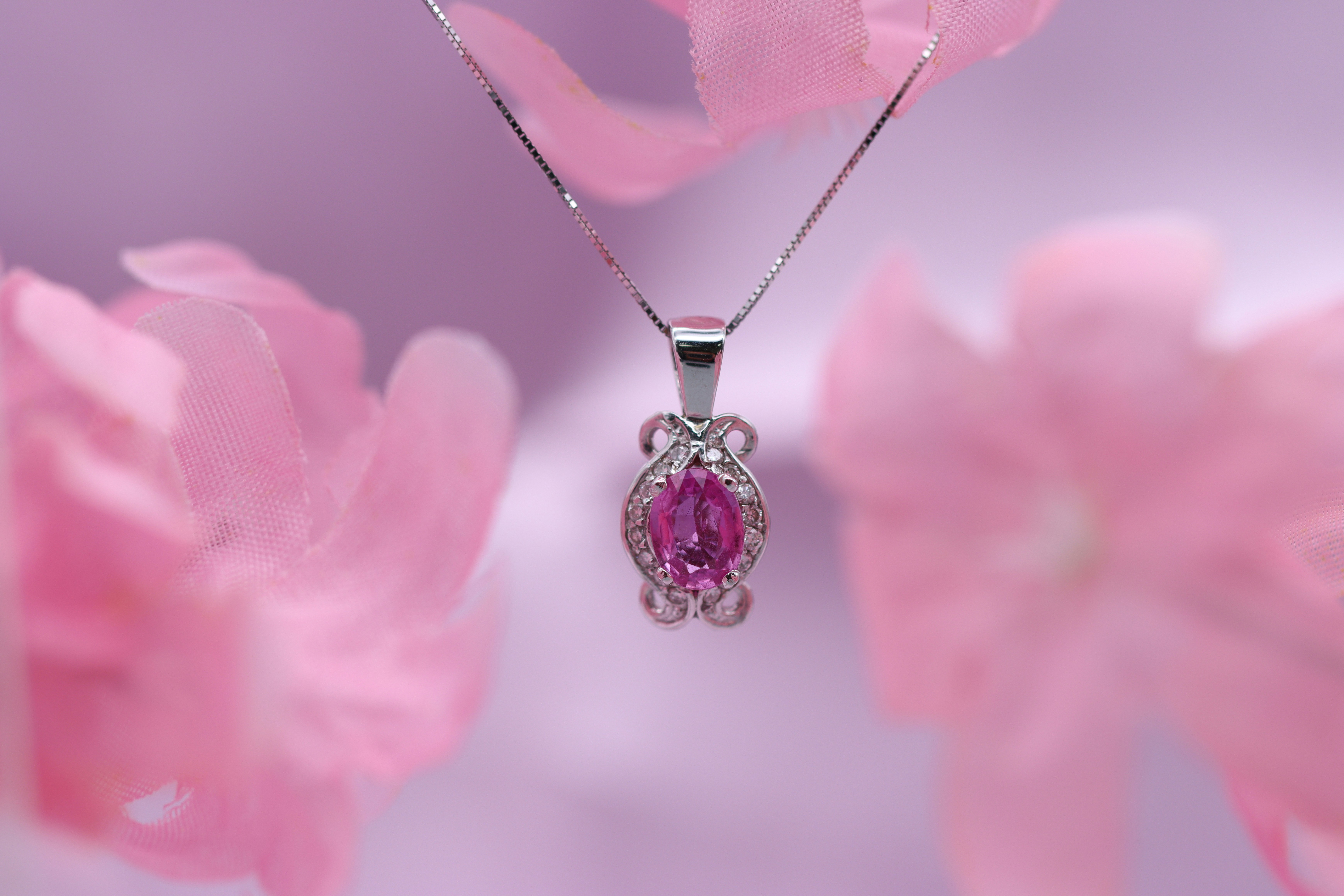 A pink gemstone pendant suspended against a backdrop of soft pink flowers, showcasing intricate silver detailing.