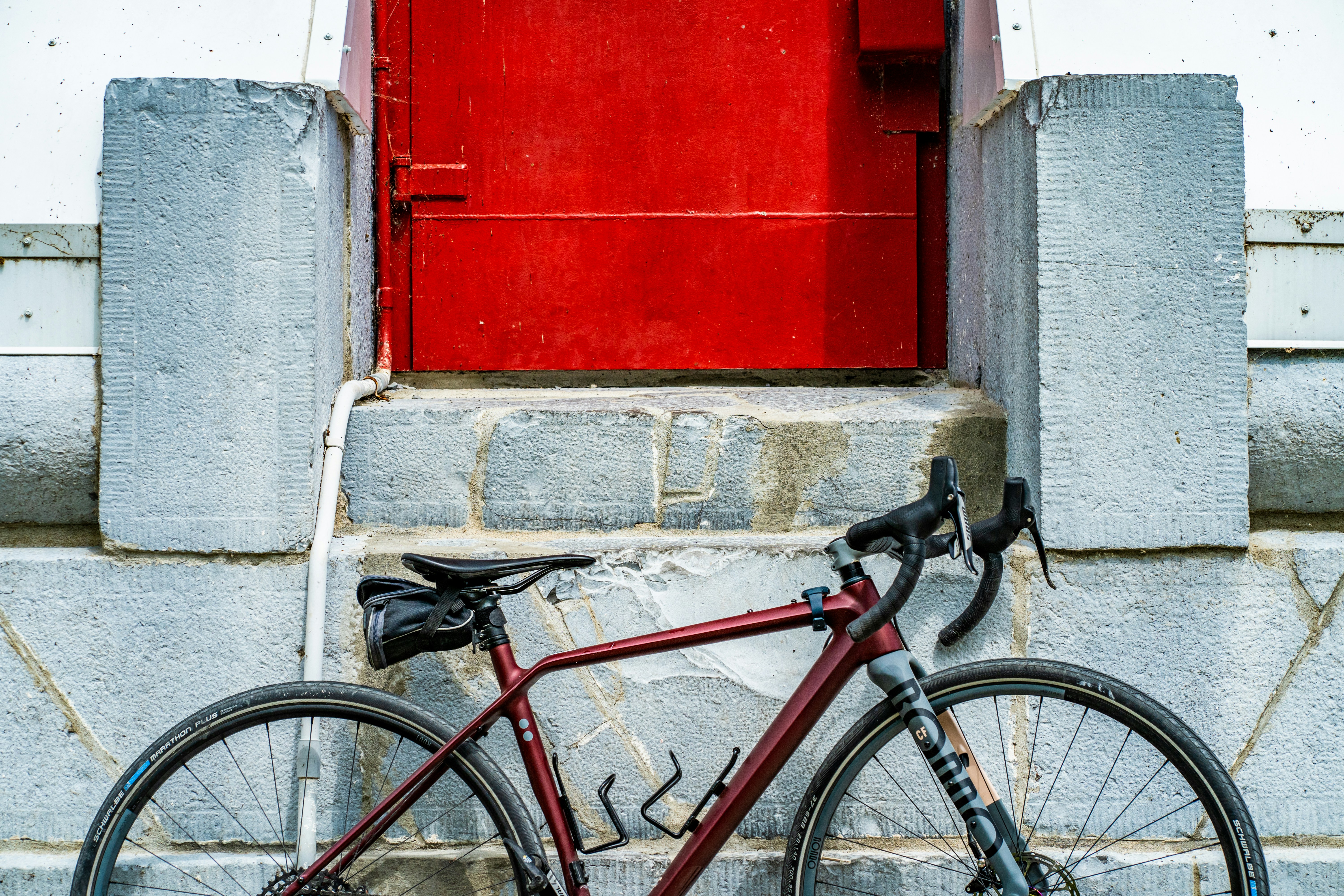 A maroon bicycle rests against a textured stone wall, leading up to a vibrant red door, showcasing urban exploration and adventure.