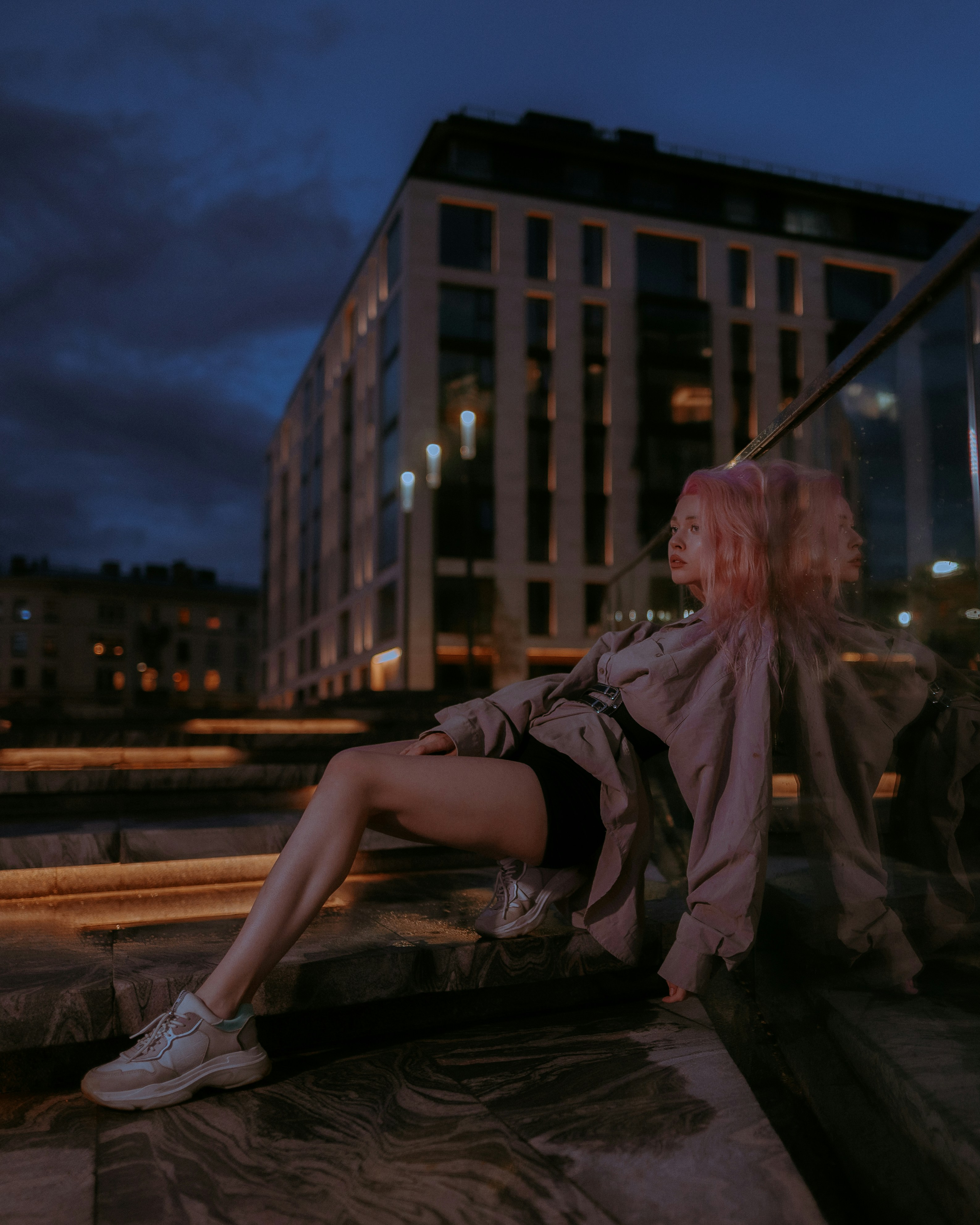 A young woman with pink hair lounges on illuminated steps, framed by modern architecture under a twilight sky.