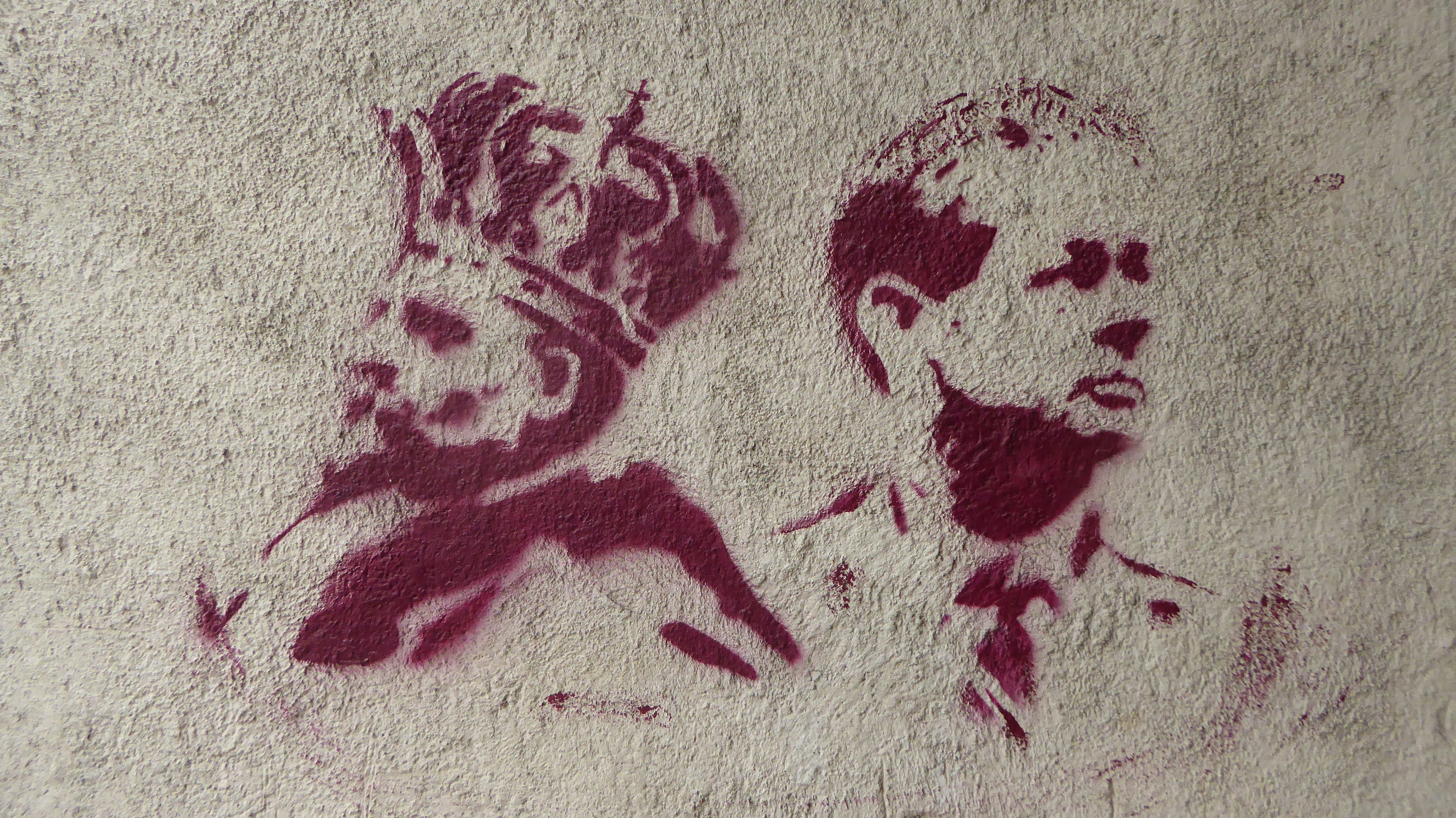 A maroon graffiti stencil painting on a rough gray wall, depicting two men's portraits; the left figure wears a crown while the right is shown in profile.